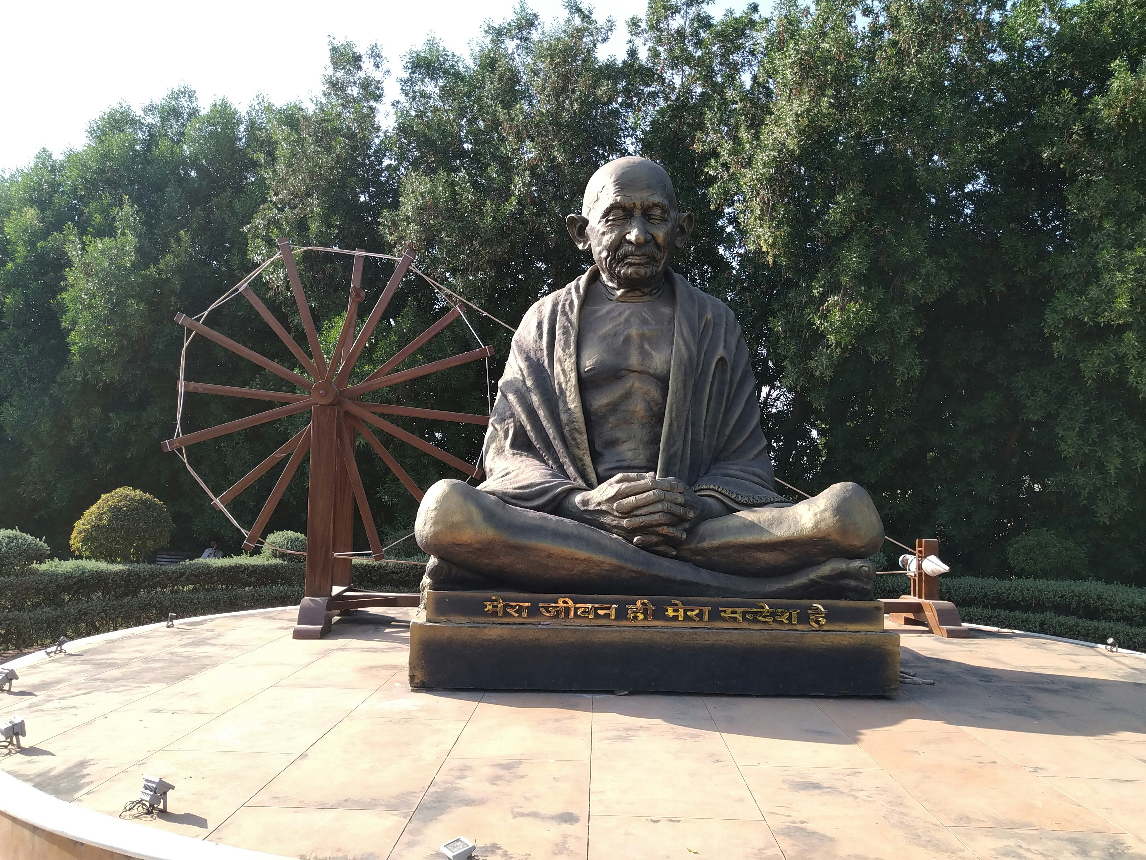 Bronze statue of Mahatma Gandhi seated in meditation, with a spinning wheel in the background, symbolizing self-reliance and peace.