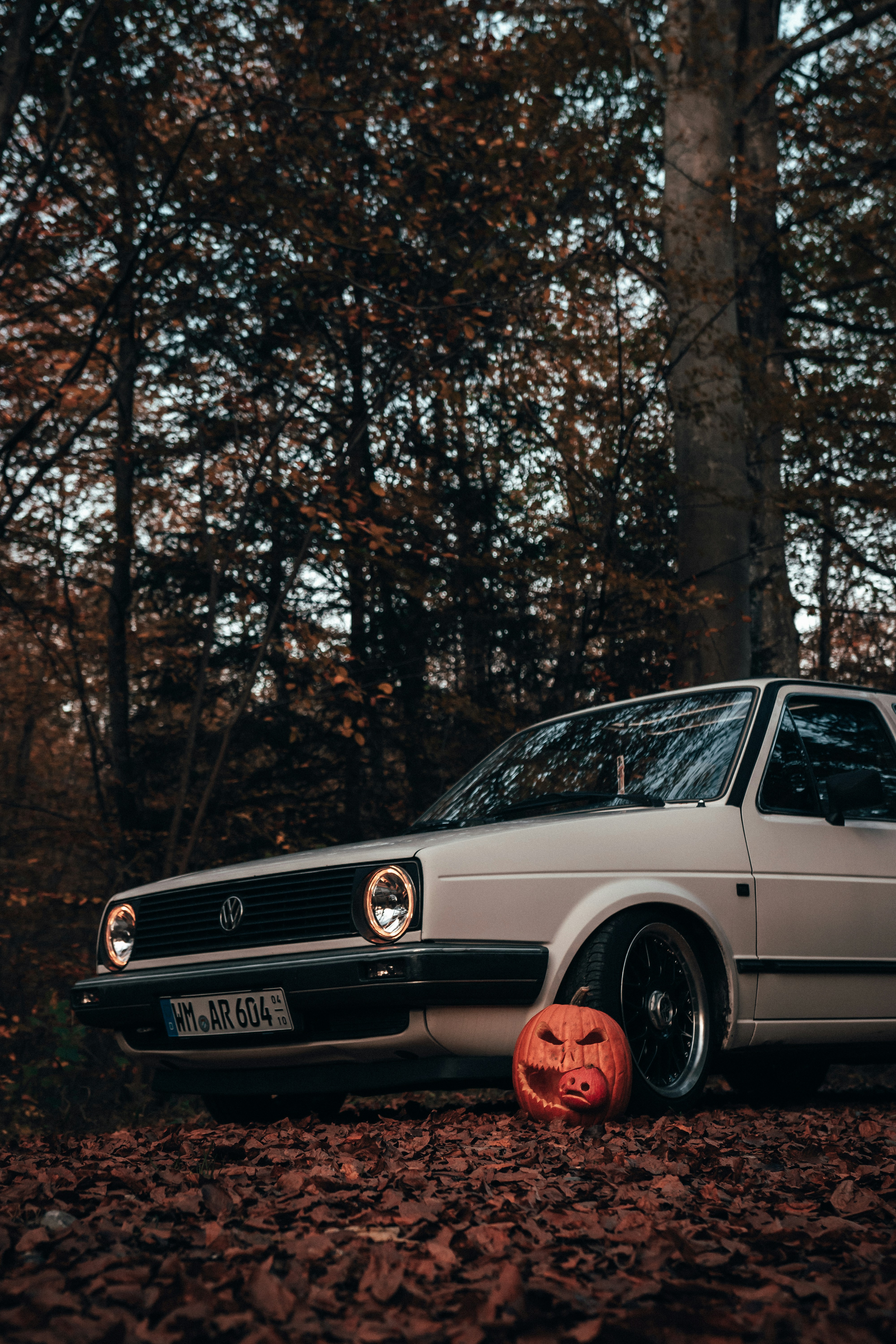A vintage white Volkswagen parked in a leaf-strewn forest with a carved pumpkin beside the front wheel, captured in moody autumn light.