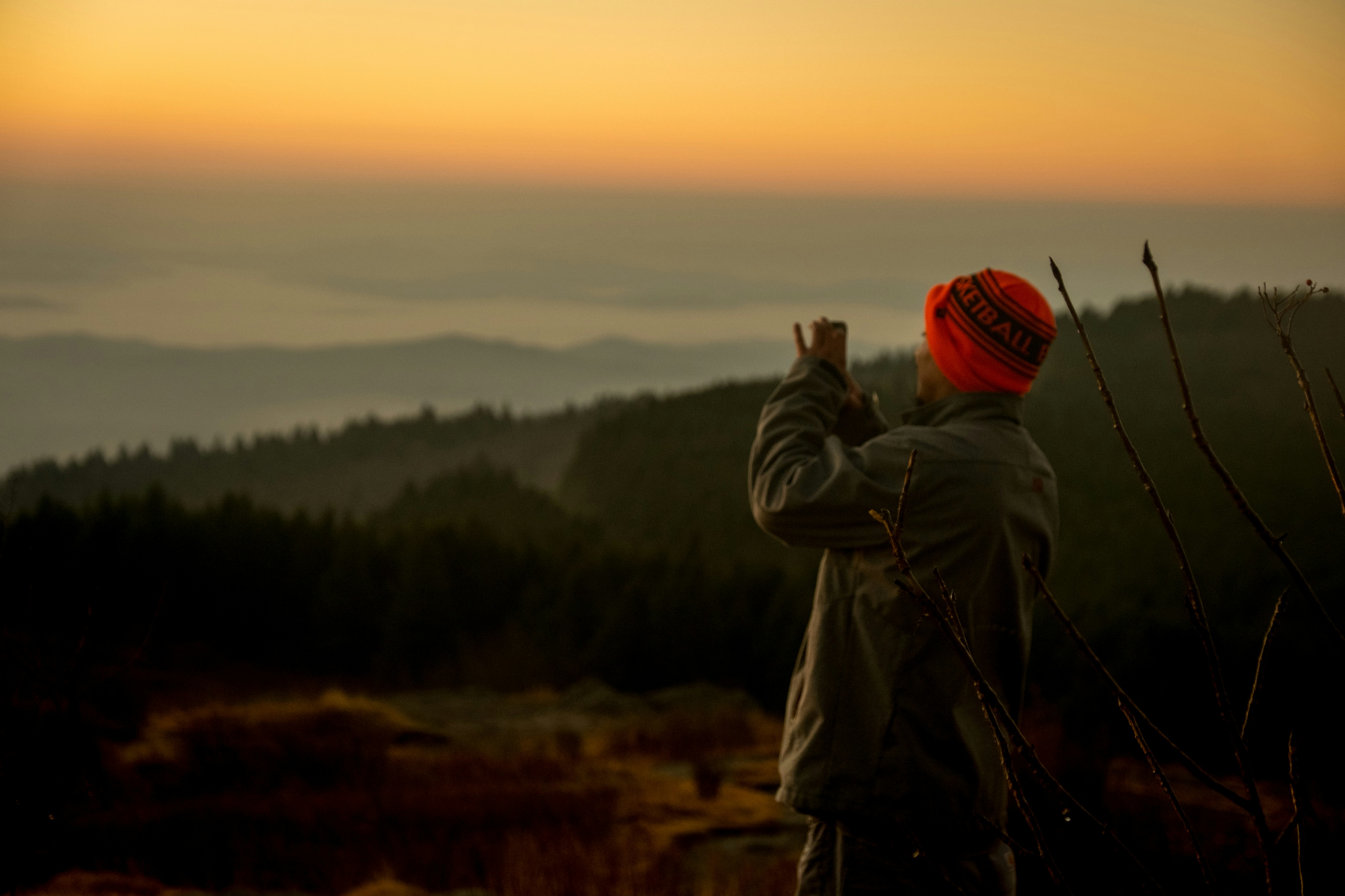 Person in a red beanie capturing the sunrise with a smartphone atop a misty mountain landscape.