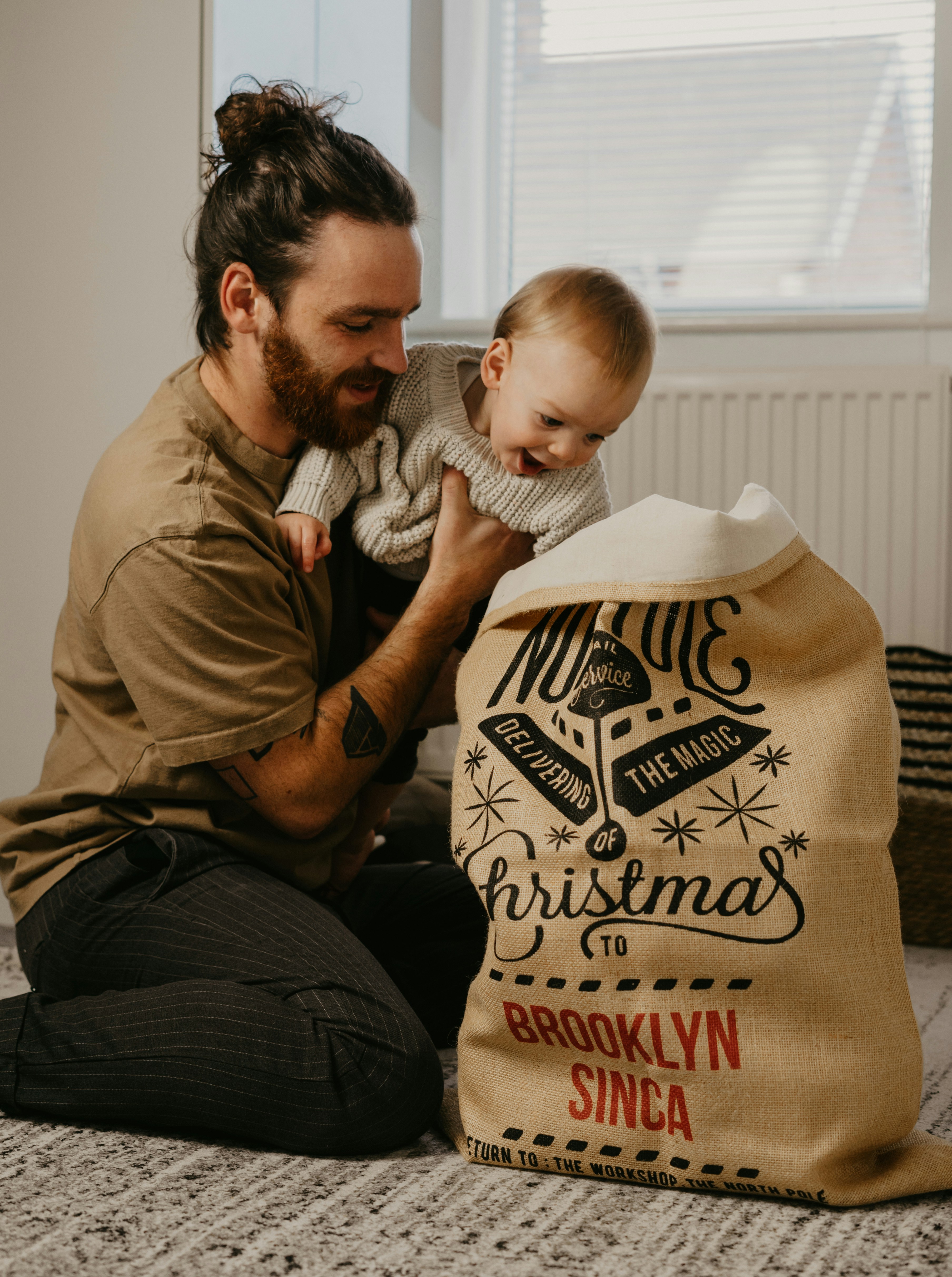 Man holding a smiling baby next to a large Christmas-themed sack indoors.