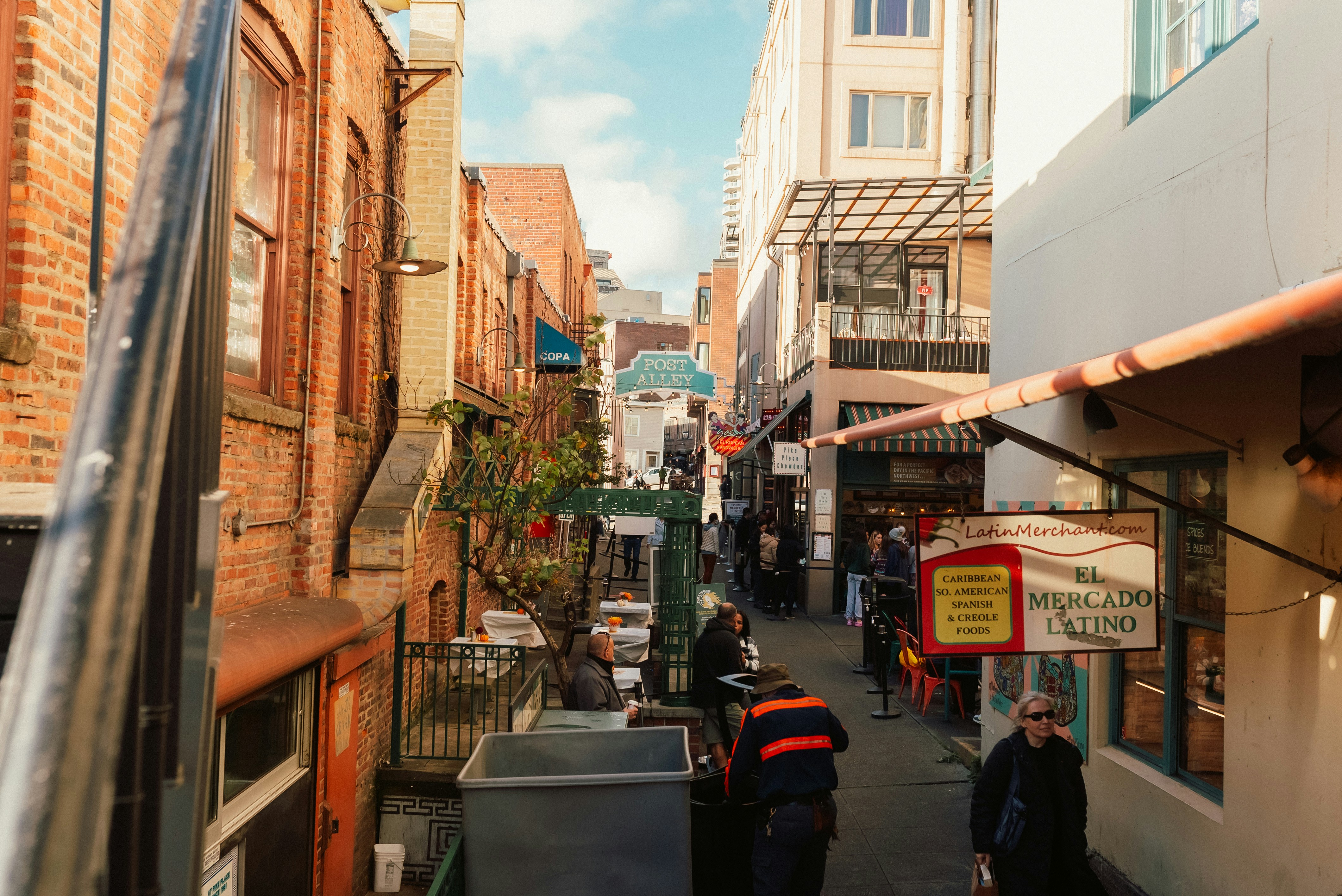 Bustling alleyway with brick buildings, shops, and people under a clear blue sky.