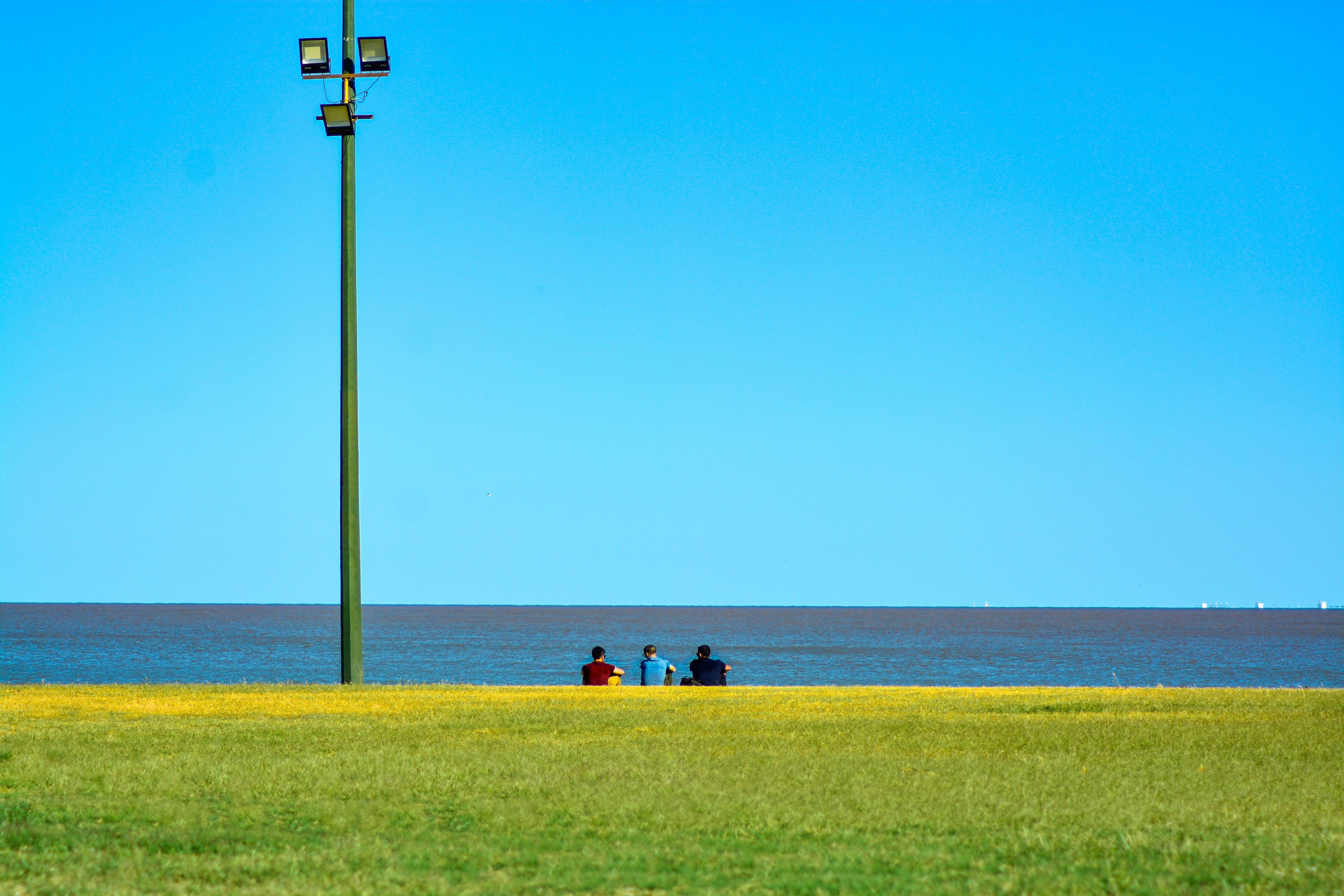 Two people seated on a bench near a tall lamp post, overlooking a vast expanse of water under a clear blue sky.