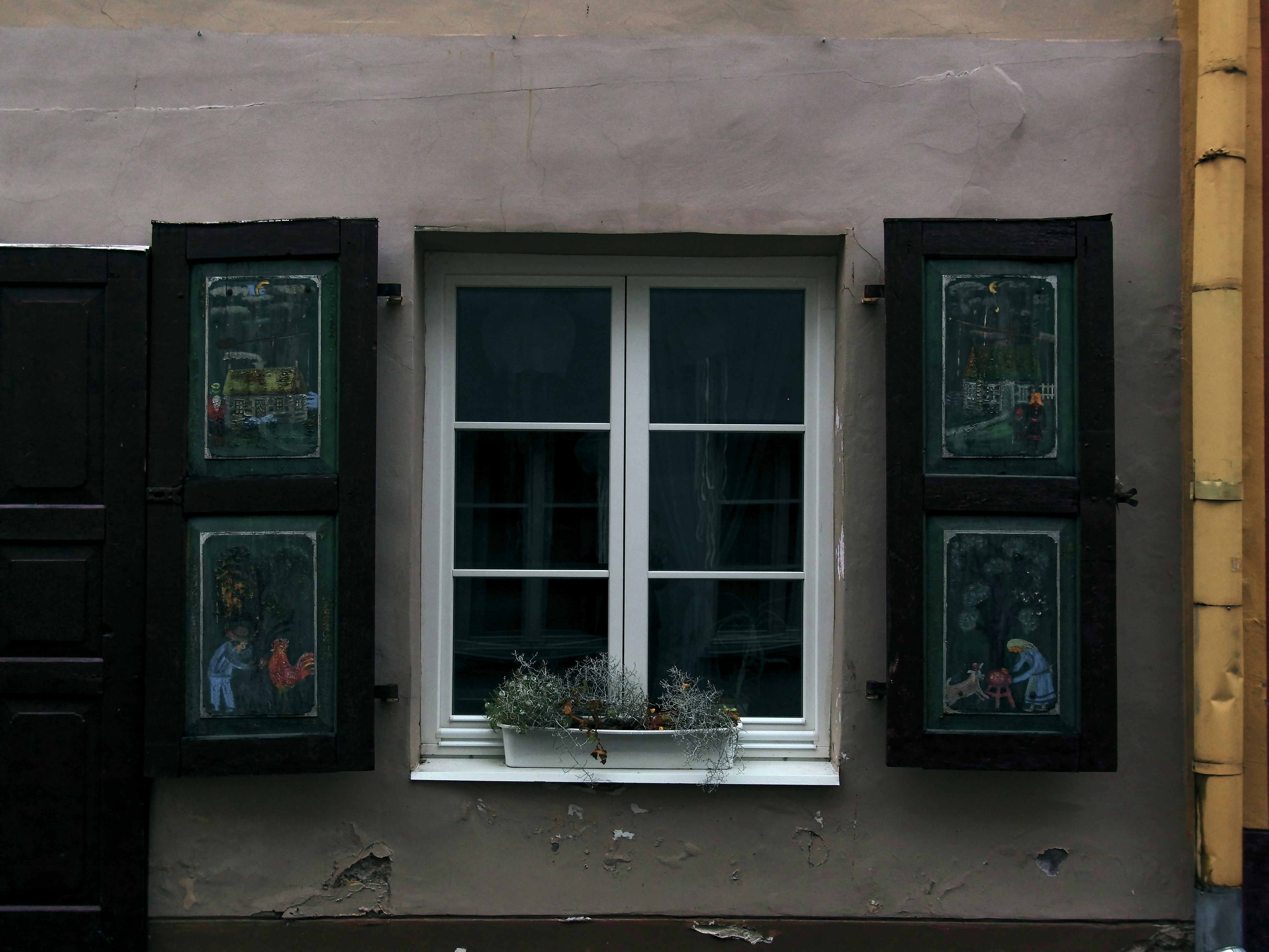 Centered white-framed window with a small flower box, flanked by decorative dark shutters on a weathered wall. Muted tones and subtle wear add character to this quiet urban detail.