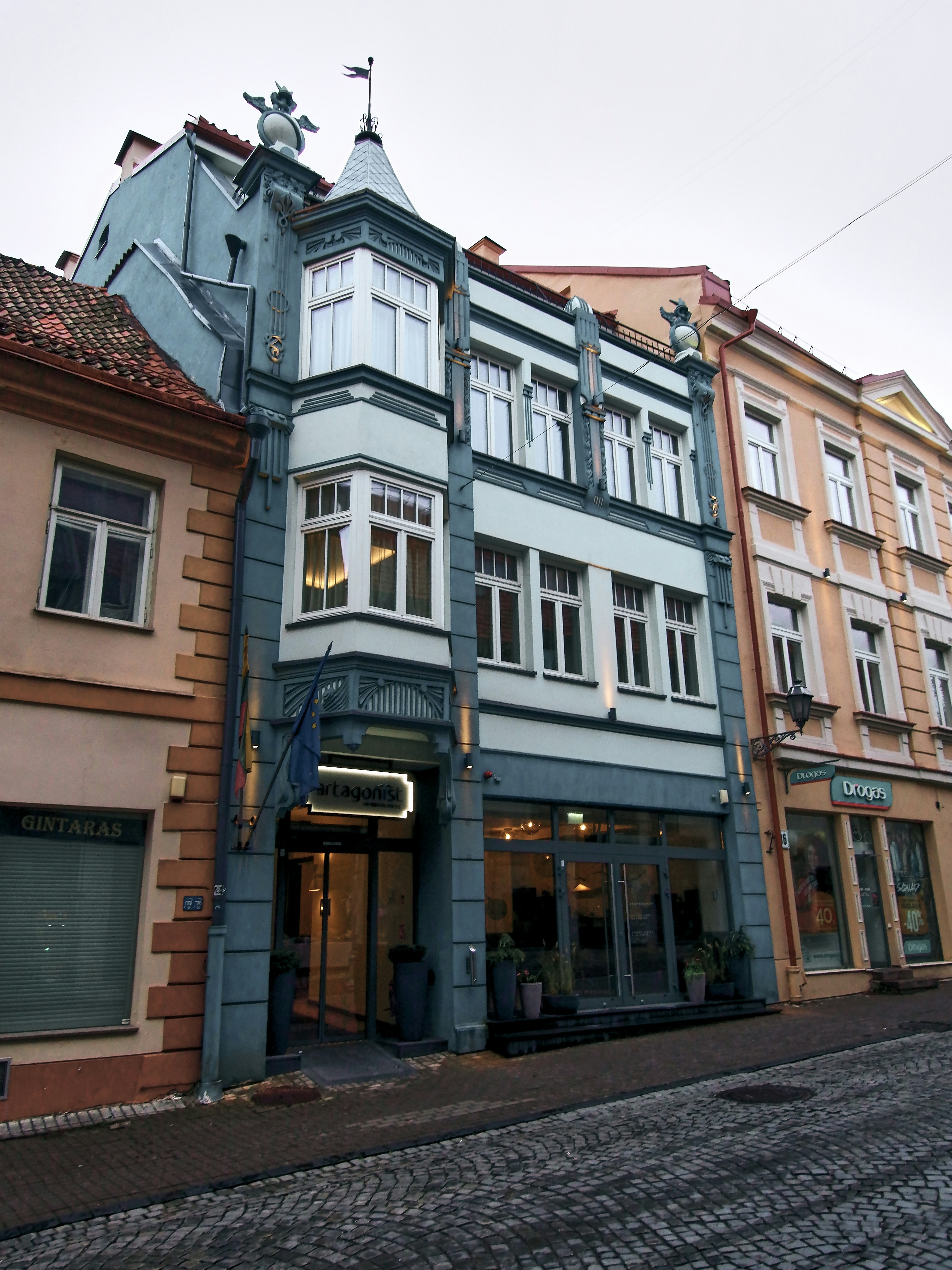 Blue, multi-window townhouse anchors a cobblestone street, flanked by pastel storefronts. The scene captures a quiet European urban facade at street level with a warmly lit entrance.