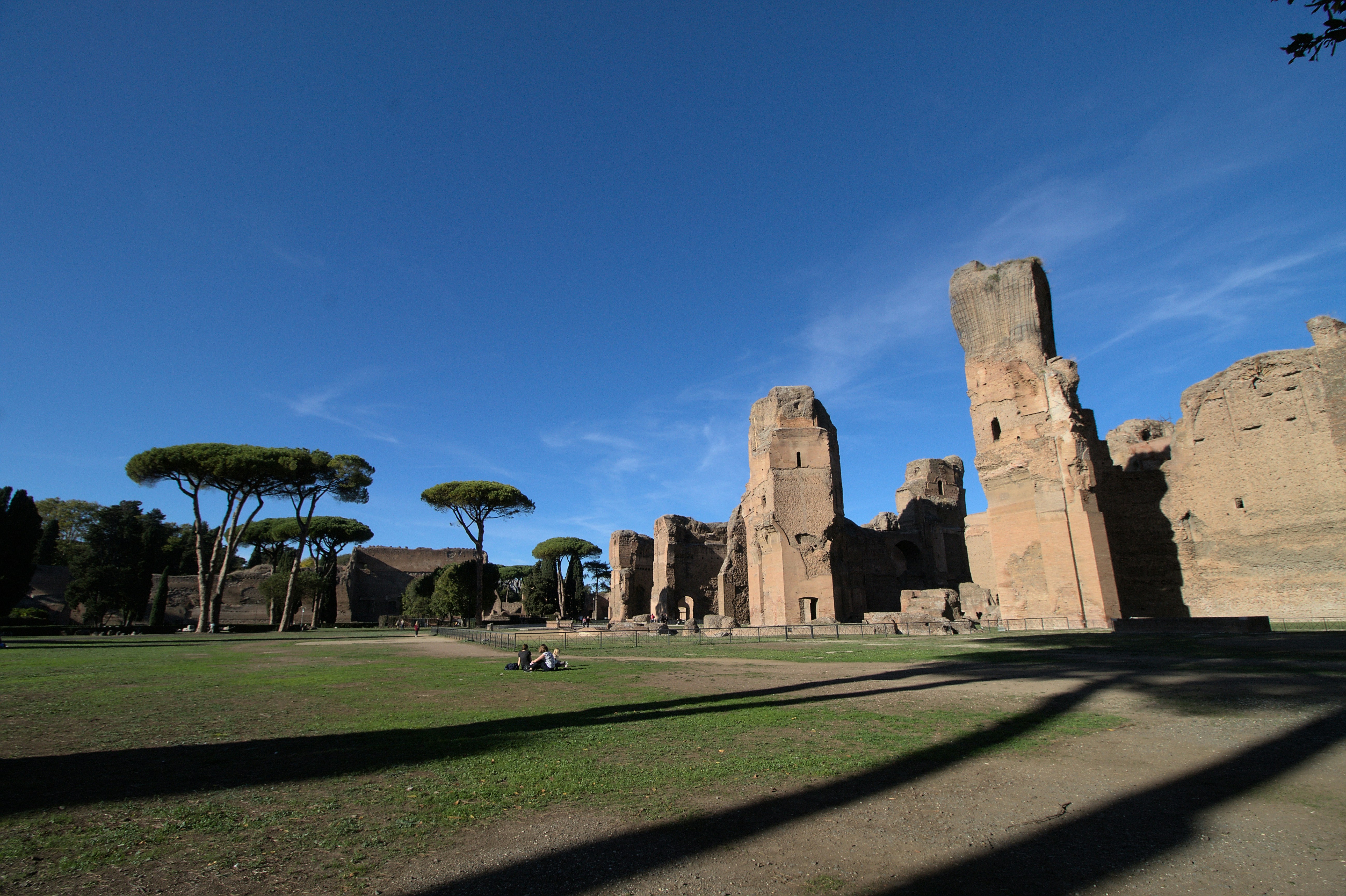 Ruins of an ancient structure with tall stone walls and scattered trees under a bright blue sky.