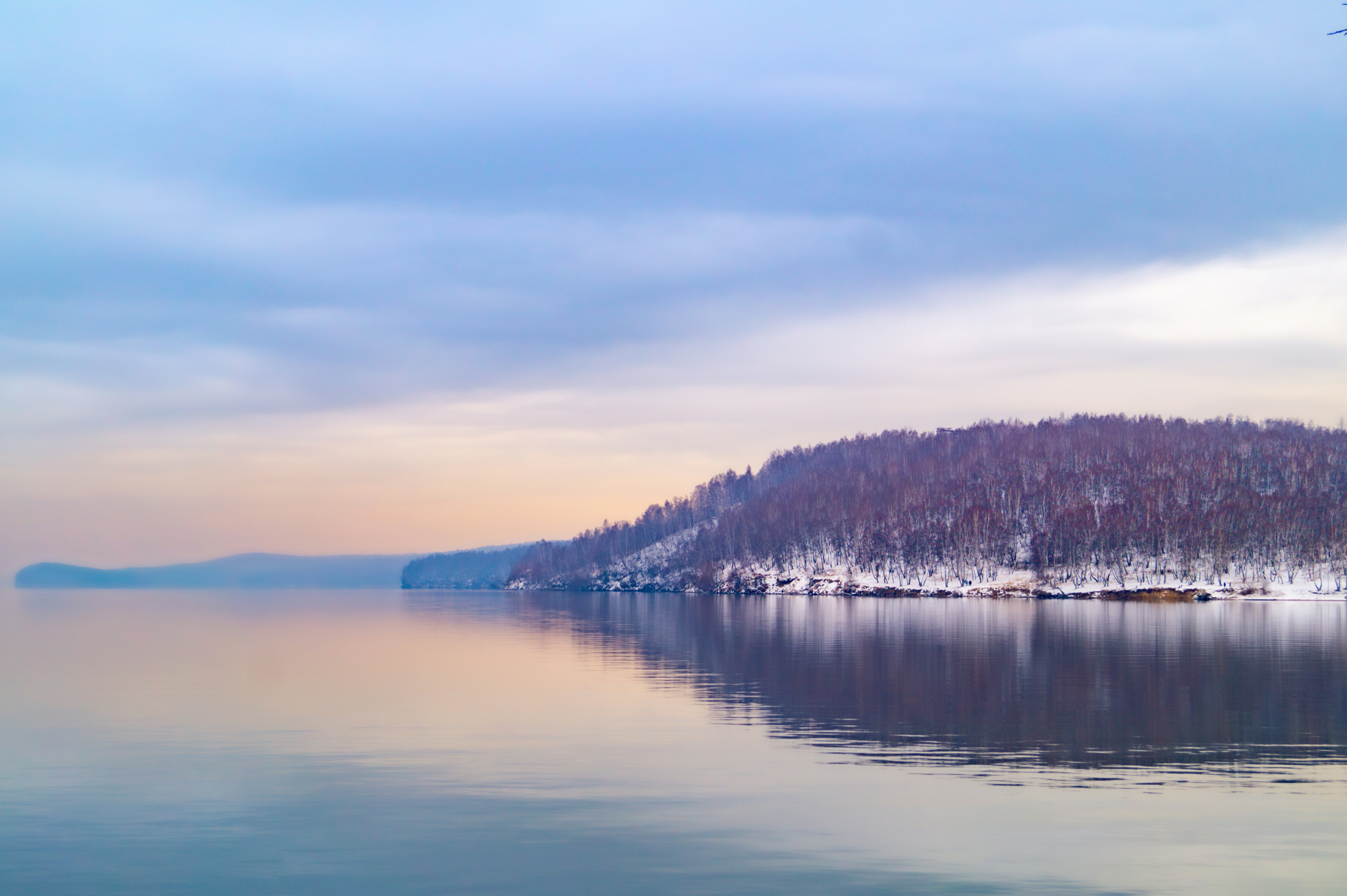 Serene winter landscape featuring a calm lake reflecting a gentle pastel sky and a snow-covered shoreline.
