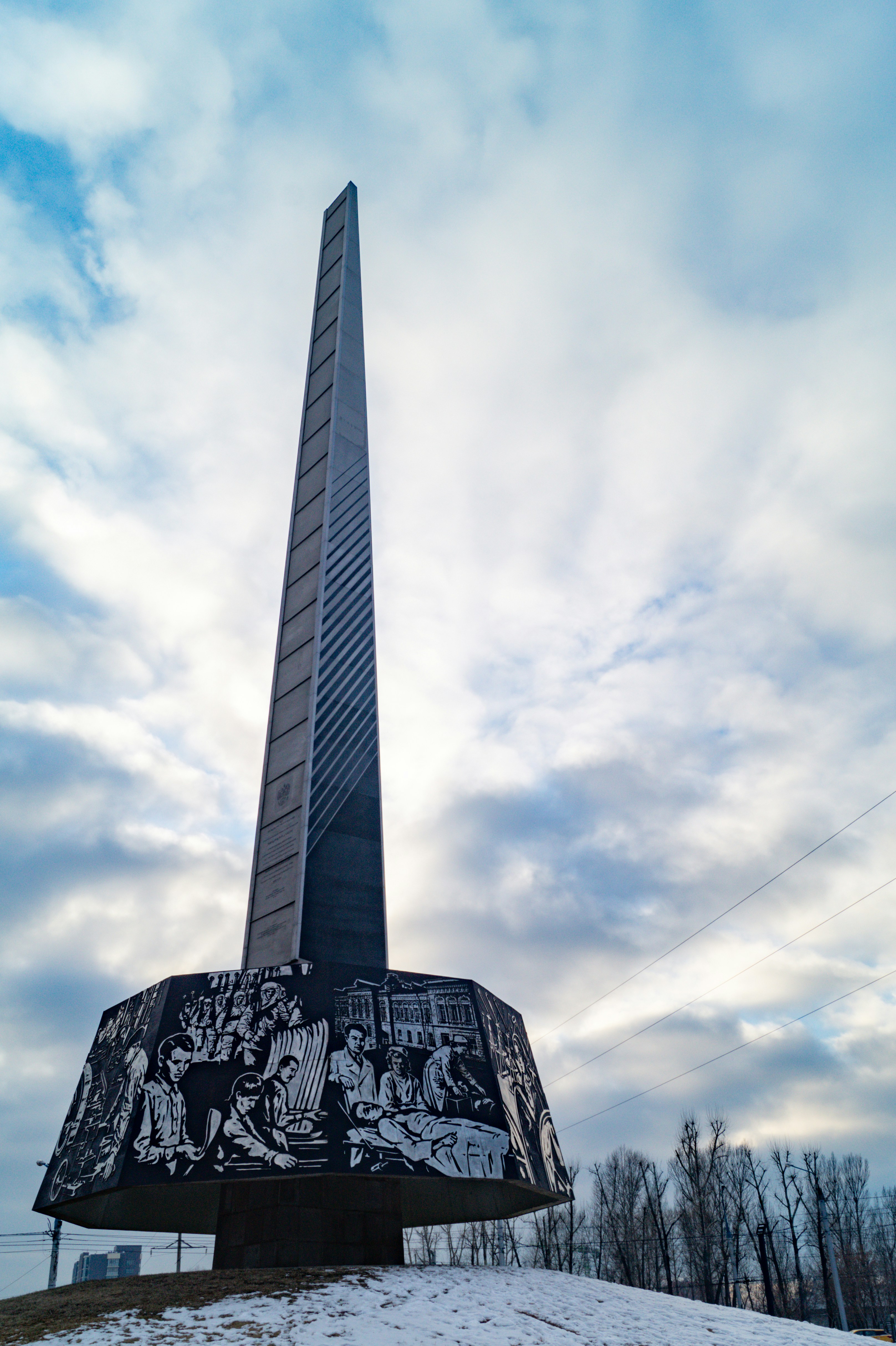 Sleek, towering monument featuring intricate black and white illustrations depicting historical scenes at its base, set against a cloudy sky. The structure symbolizes resilience and remembrance.