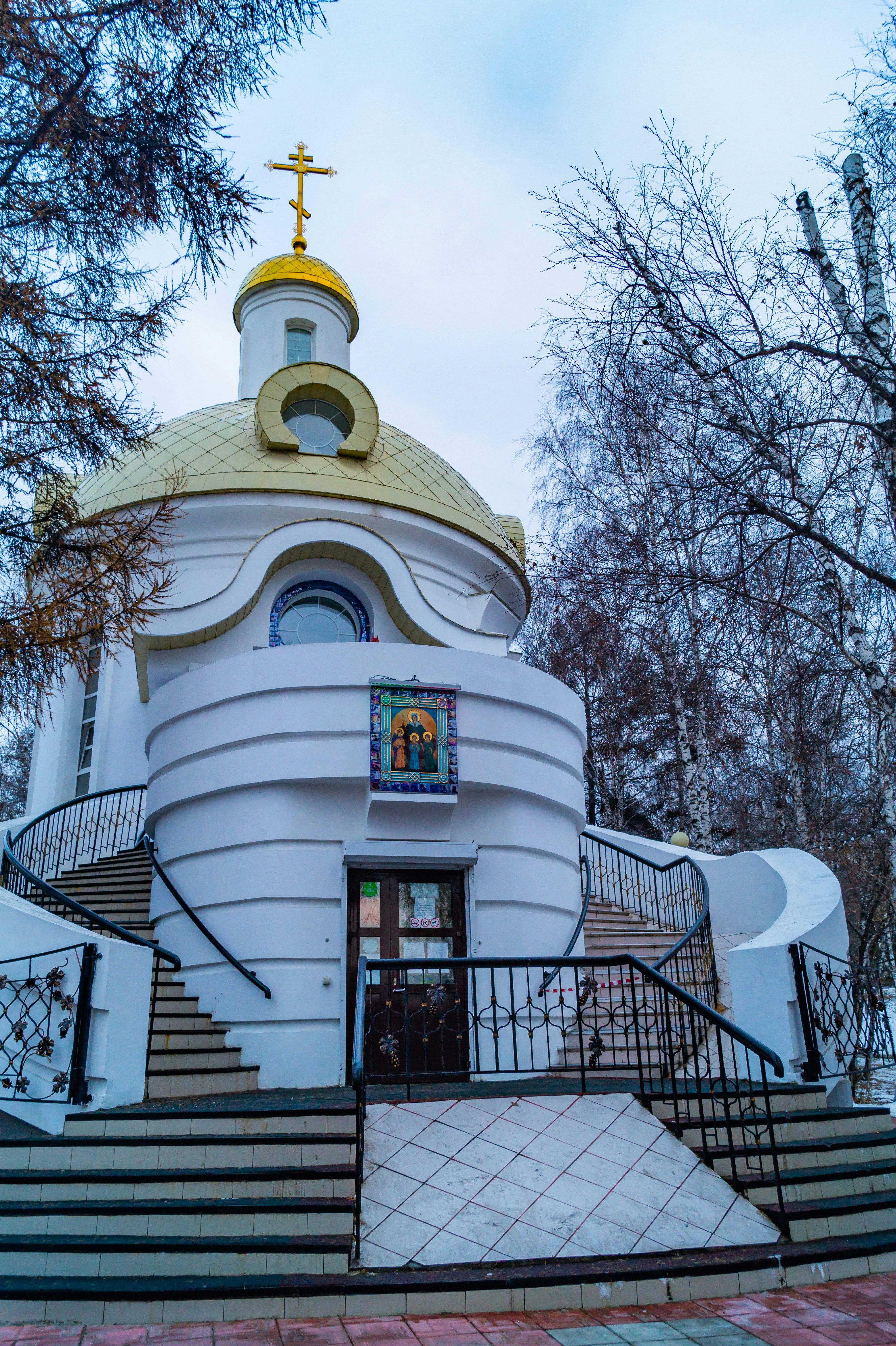 A modern church featuring a golden dome and intricate architectural details, surrounded by bare trees under a cloudy sky.