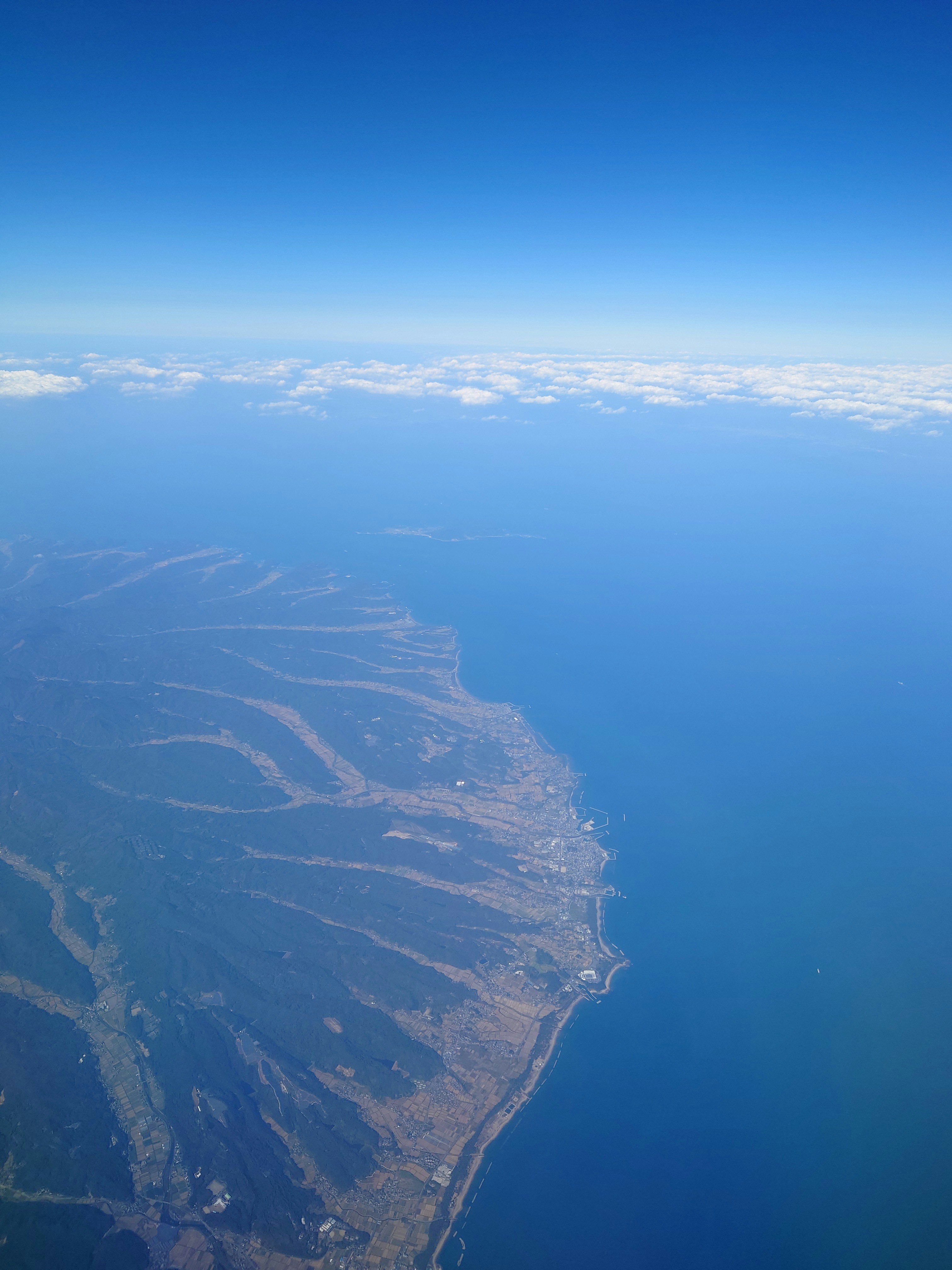 Aerial view showcasing intricate coastal lines and land formations meeting the ocean. The vast blue sky complements the earthy tones of the landscape.