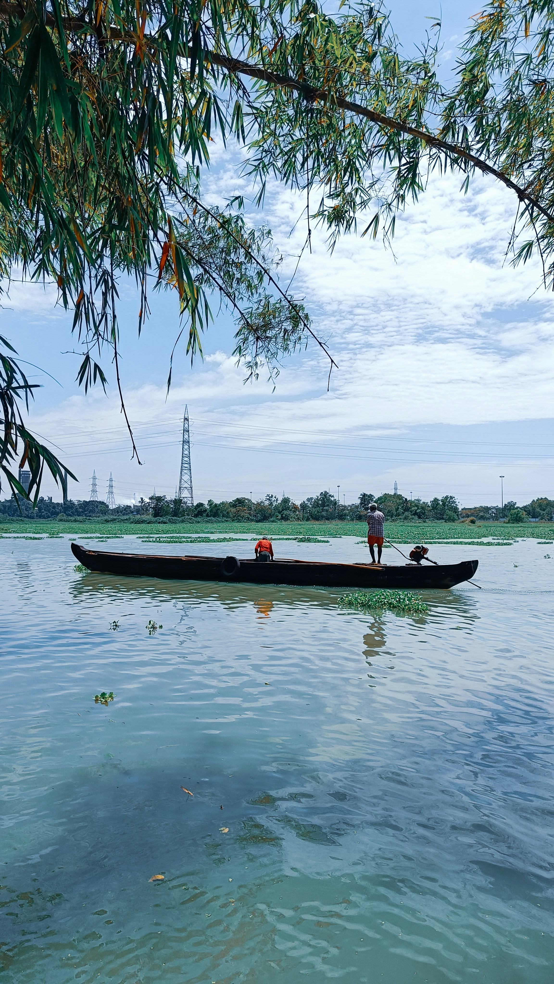 Two fishermen in a wooden boat navigate through a serene river, surrounded by lush greenery and distant power lines. The calm water reflects the vibrant sky.