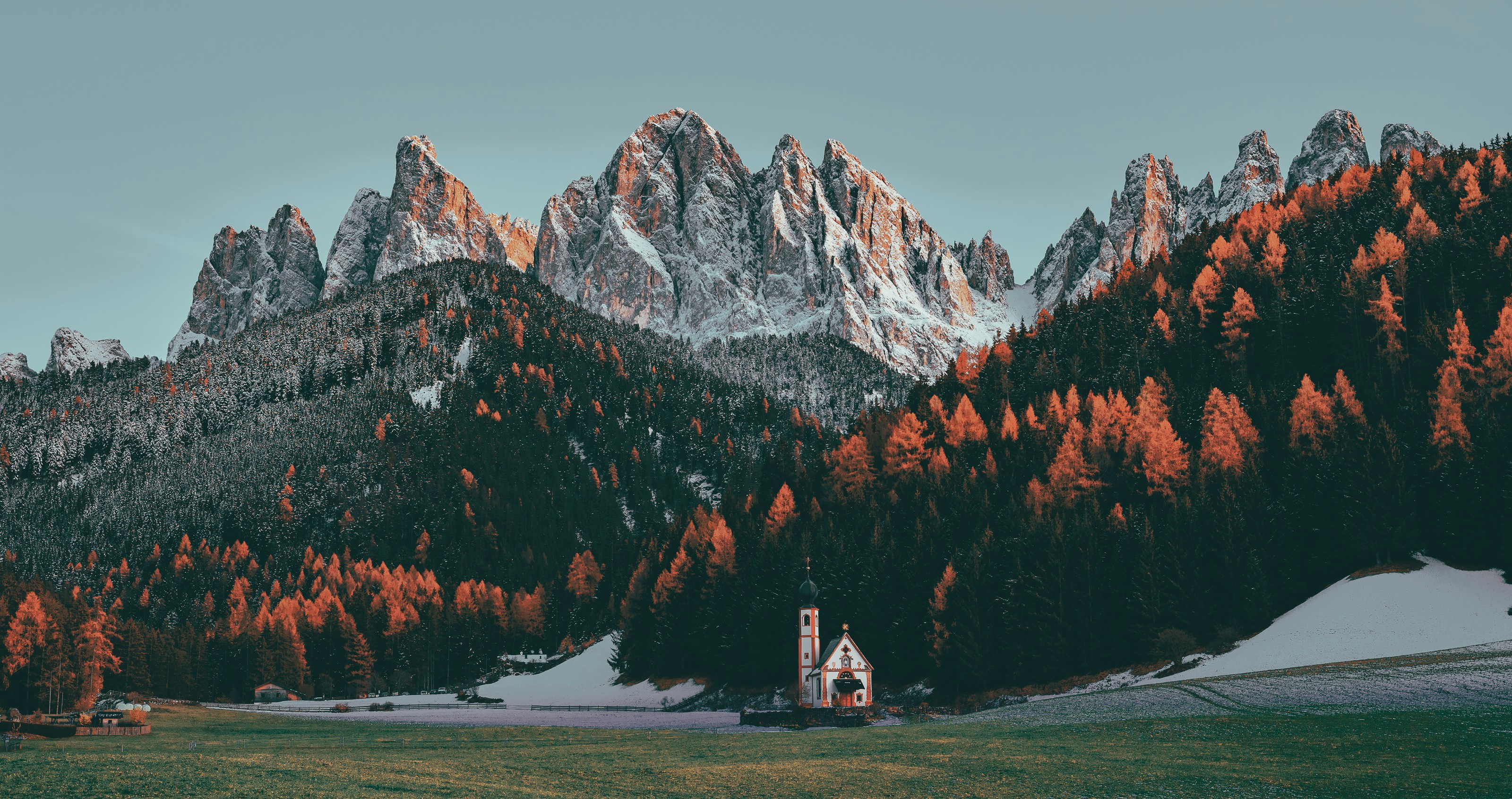 Small chapel nestled in a vibrant autumnal valley with towering jagged mountains in the background.