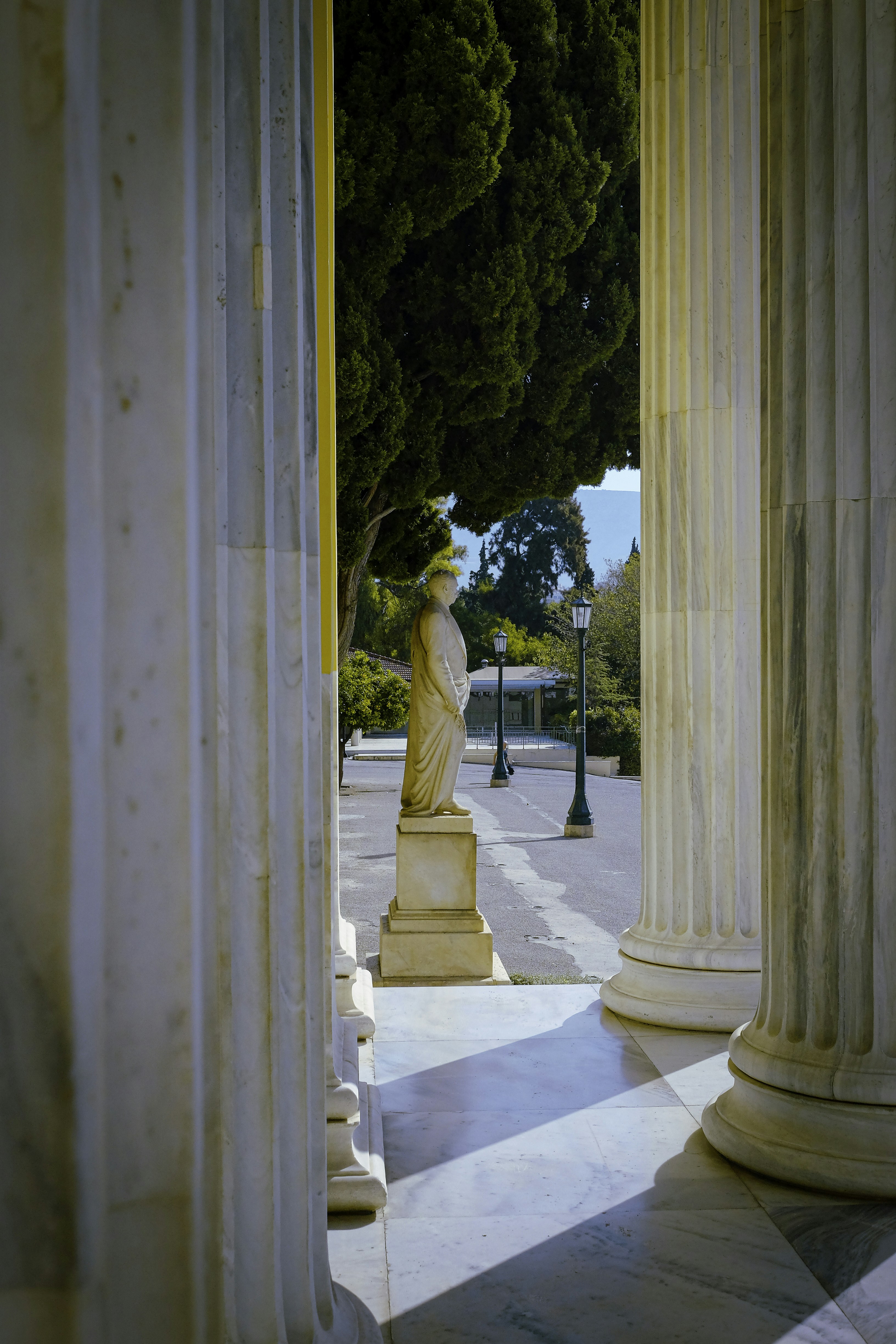 Marble statue stands between tall neoclassical columns along a sunlit walkway, with trees and a lamppost visible in the background.