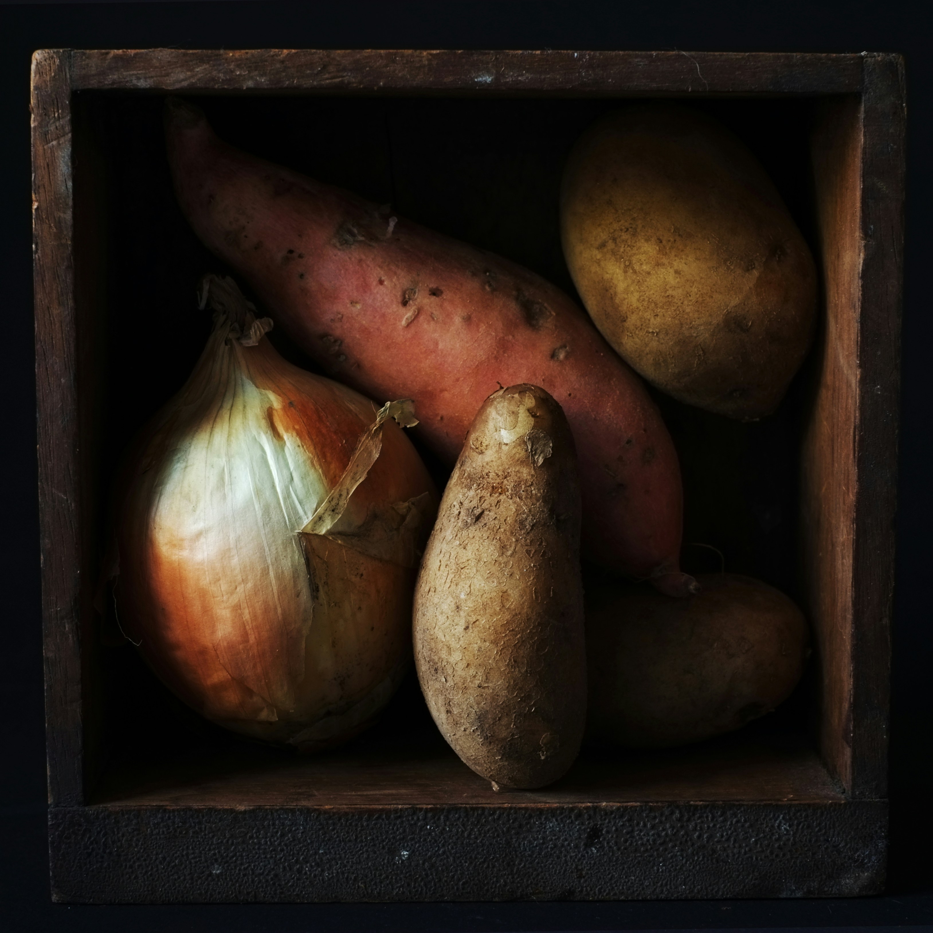 A wooden crate filled with various root vegetables, including sweet potatoes and onions, against a dark background.