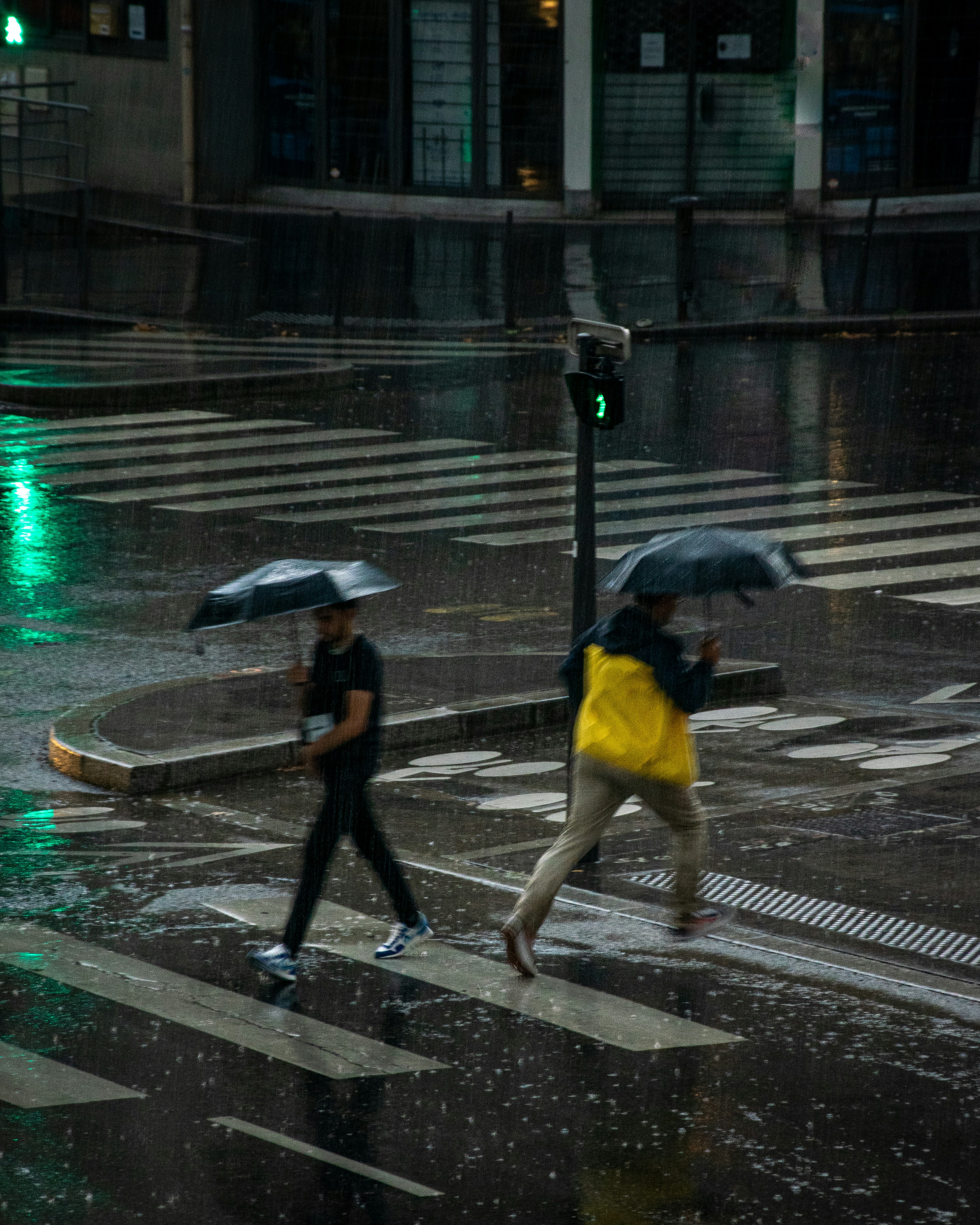Two pedestrians with umbrellas cross a rain-soaked city crosswalk at night, the yellow jacket drawing attention among neon reflections on the wet pavement.