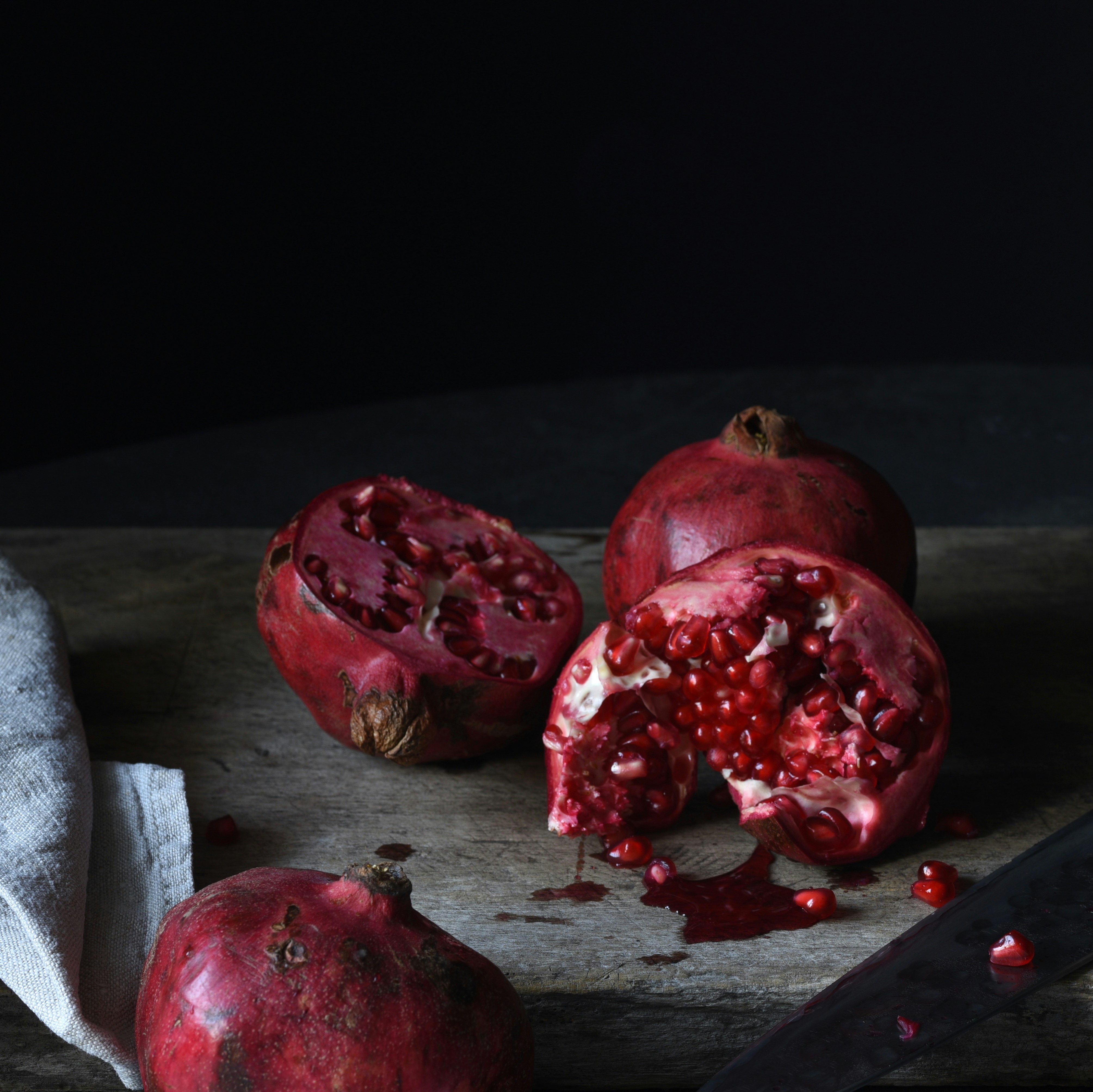 Juicy pomegranates split open on a rustic wooden surface, revealing vibrant red seeds and a hint of juice. A soft cloth adds texture to the scene.