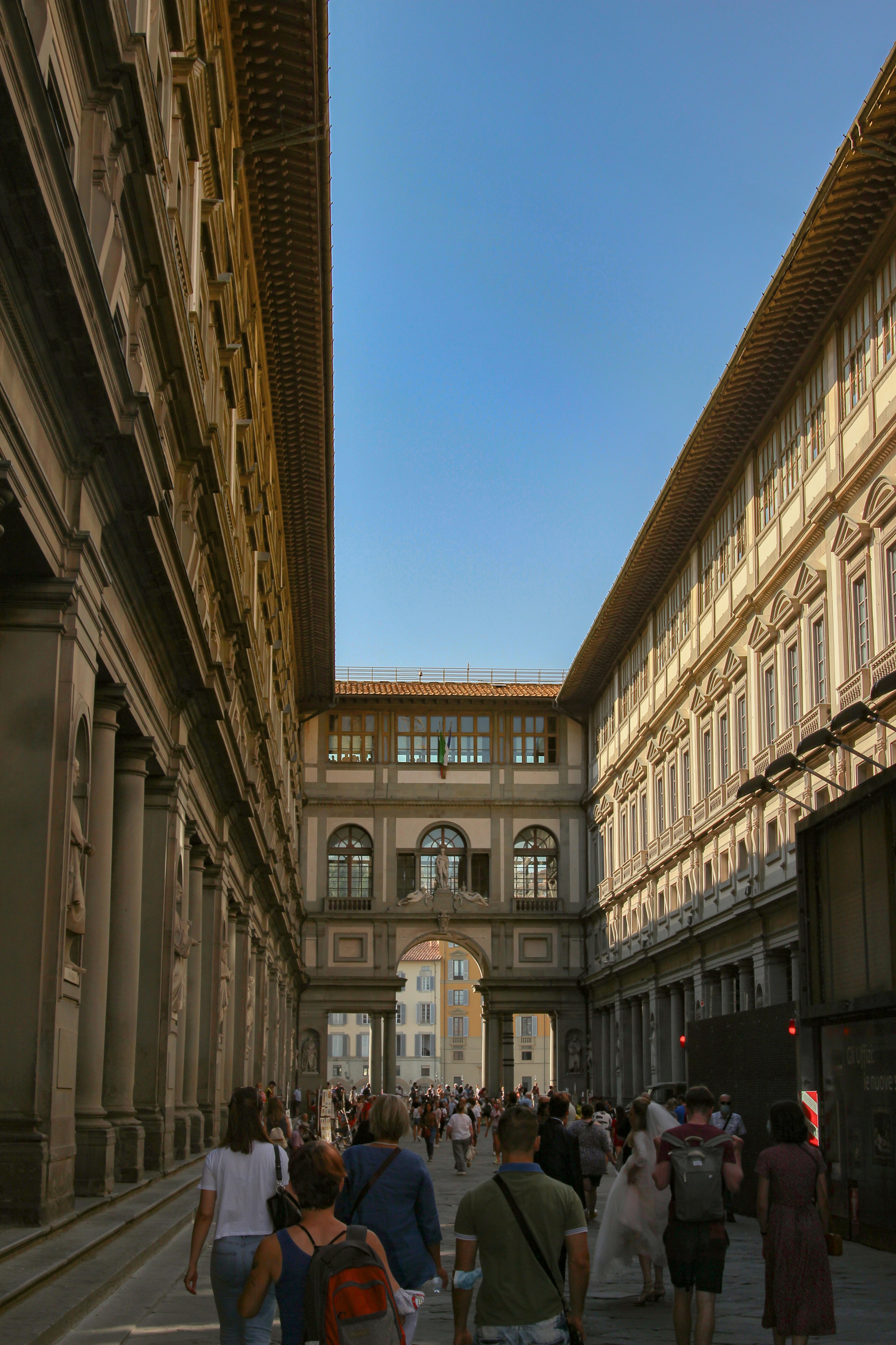 Crowd of visitors walking through historic architecture under a clear blue sky, framed by elegant buildings. The scene captures the essence of cultural exploration.