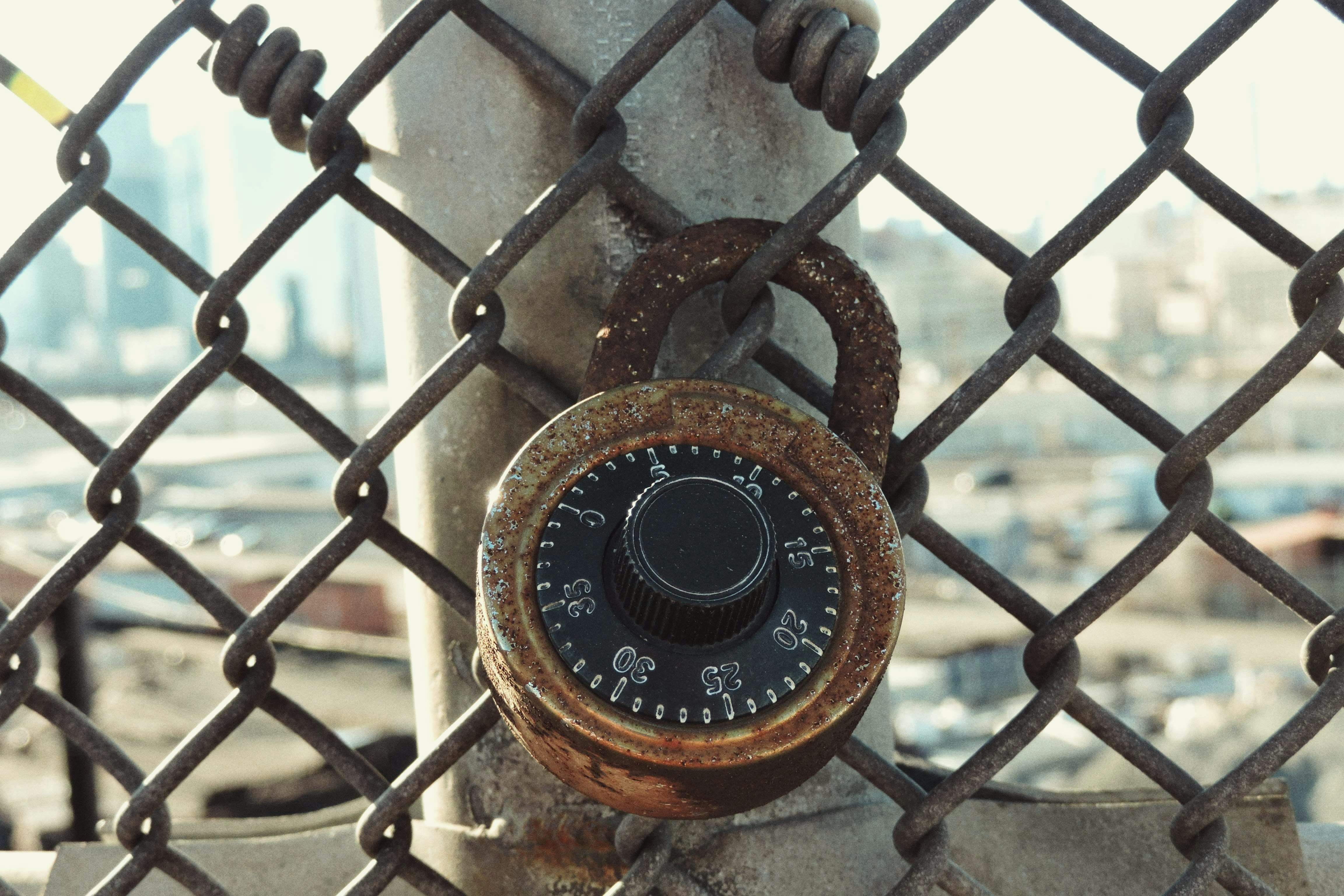 A weathered combination lock hangs on a chain-link fence, symbolizing security and forgotten stories. The background hints at an industrial setting.