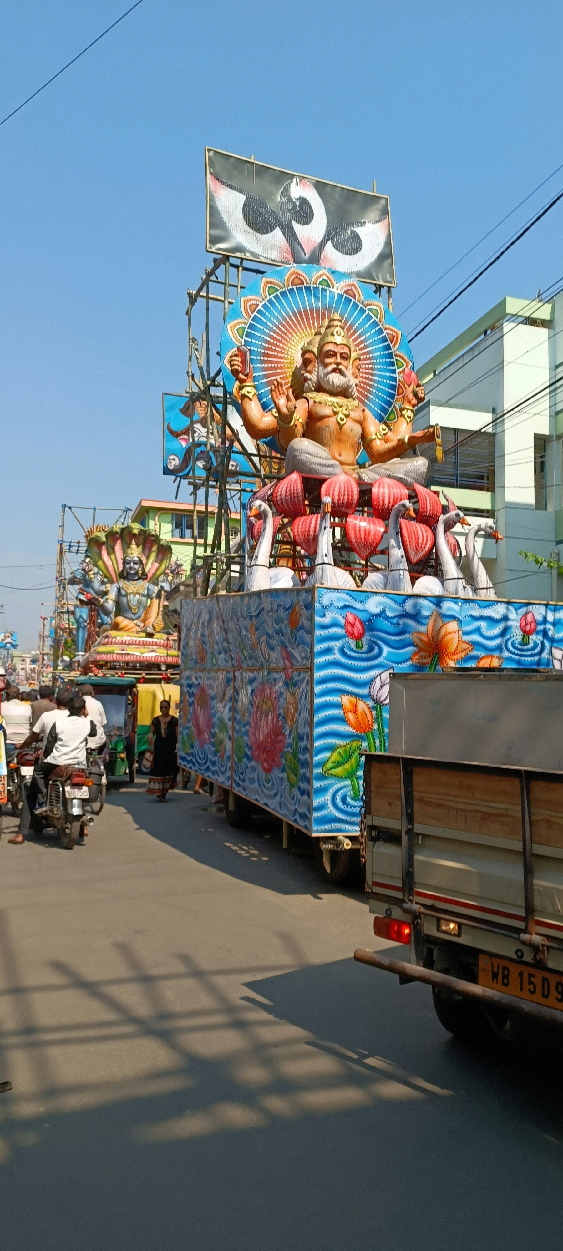 Vibrant festival float with a towering deity statue, photographed as it parades down a sunlit urban street during a colorful festival.