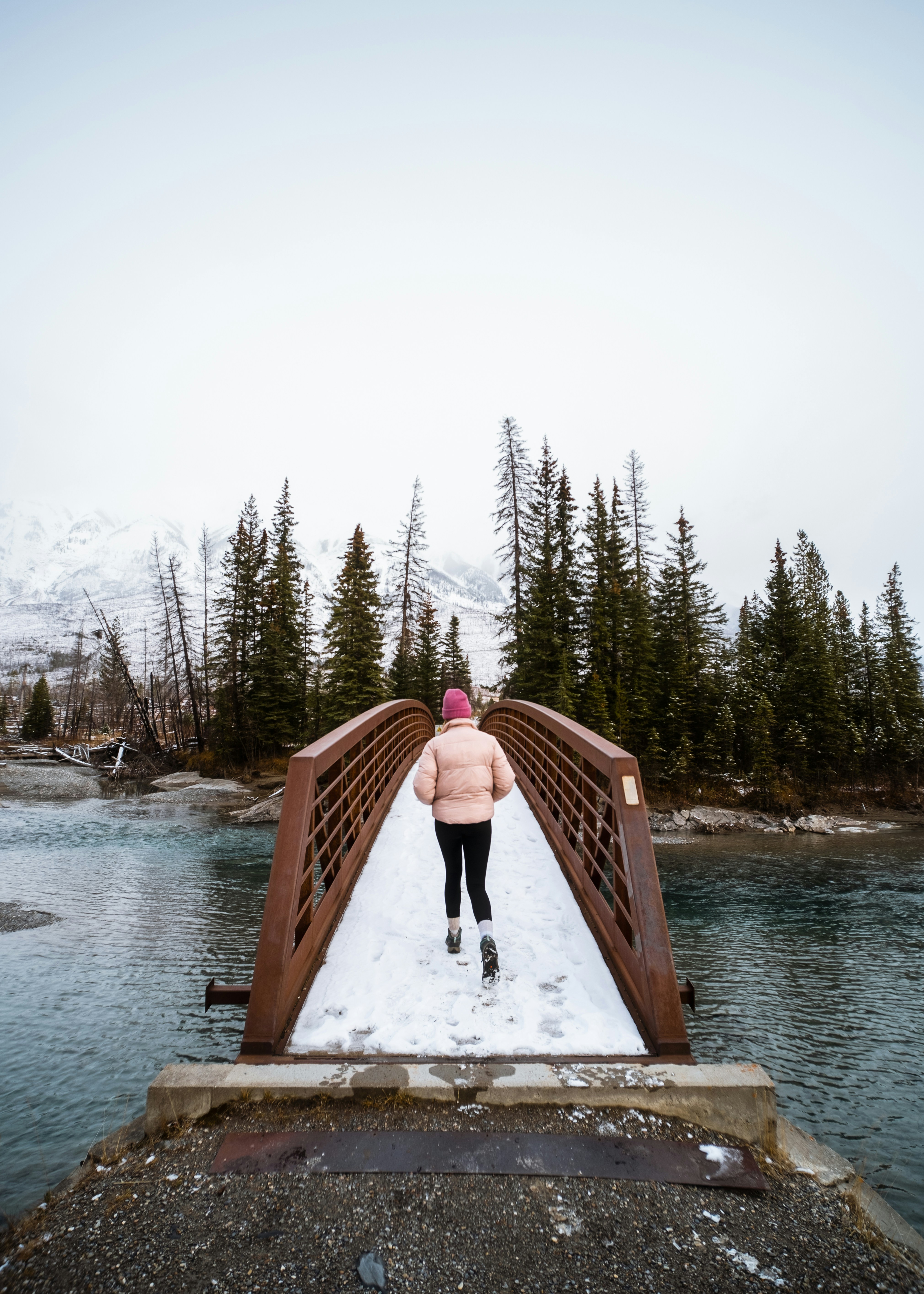 Individual in a pink jacket walking across a snow-covered bridge surrounded by evergreen trees and a river. The scene captures the essence of winter exploration.