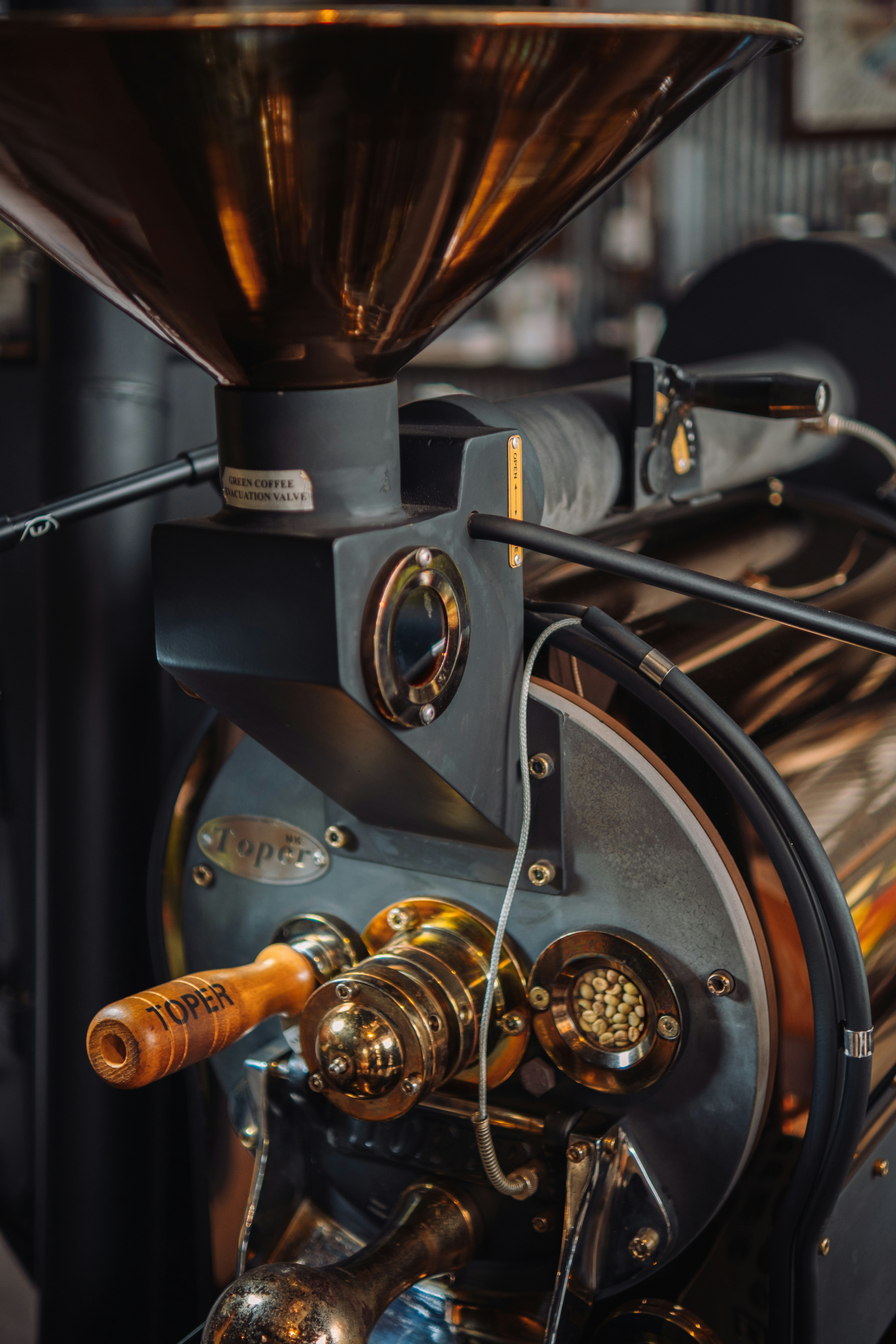 Close-up photograph of a coffee roasting machine's control assembly, including a hopper, copper fittings, and a round gauge. The image emphasizes industrial hardware and craftsmanship.