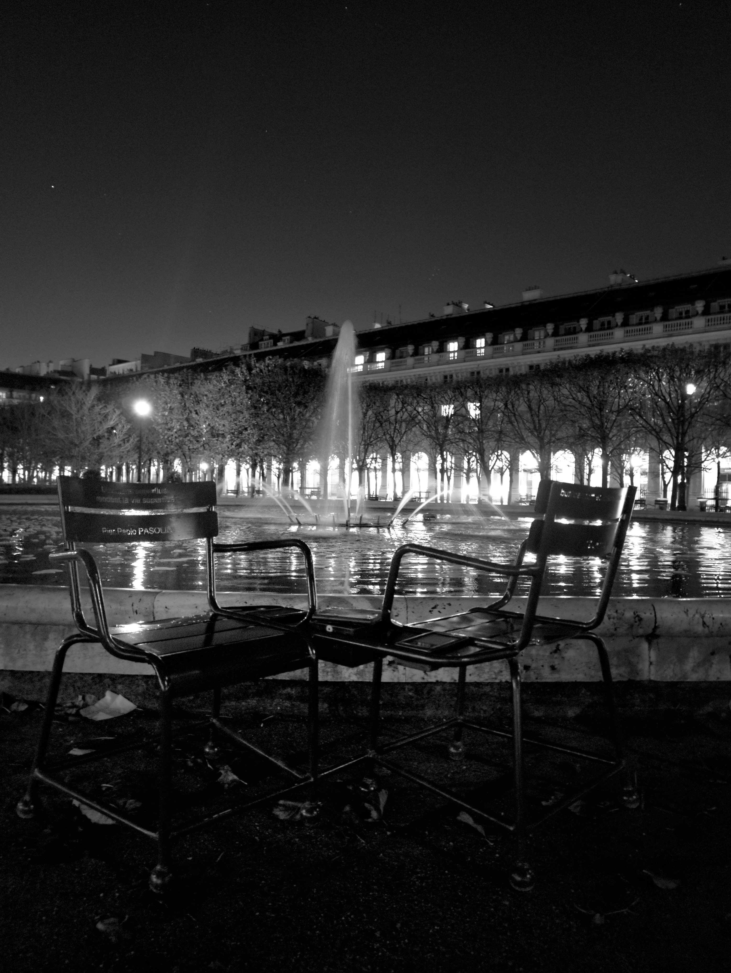 Two empty metal chairs sit beside a lit fountain in a quiet, tree-lined urban square at night. The background features illuminated arches and reflections on the water.