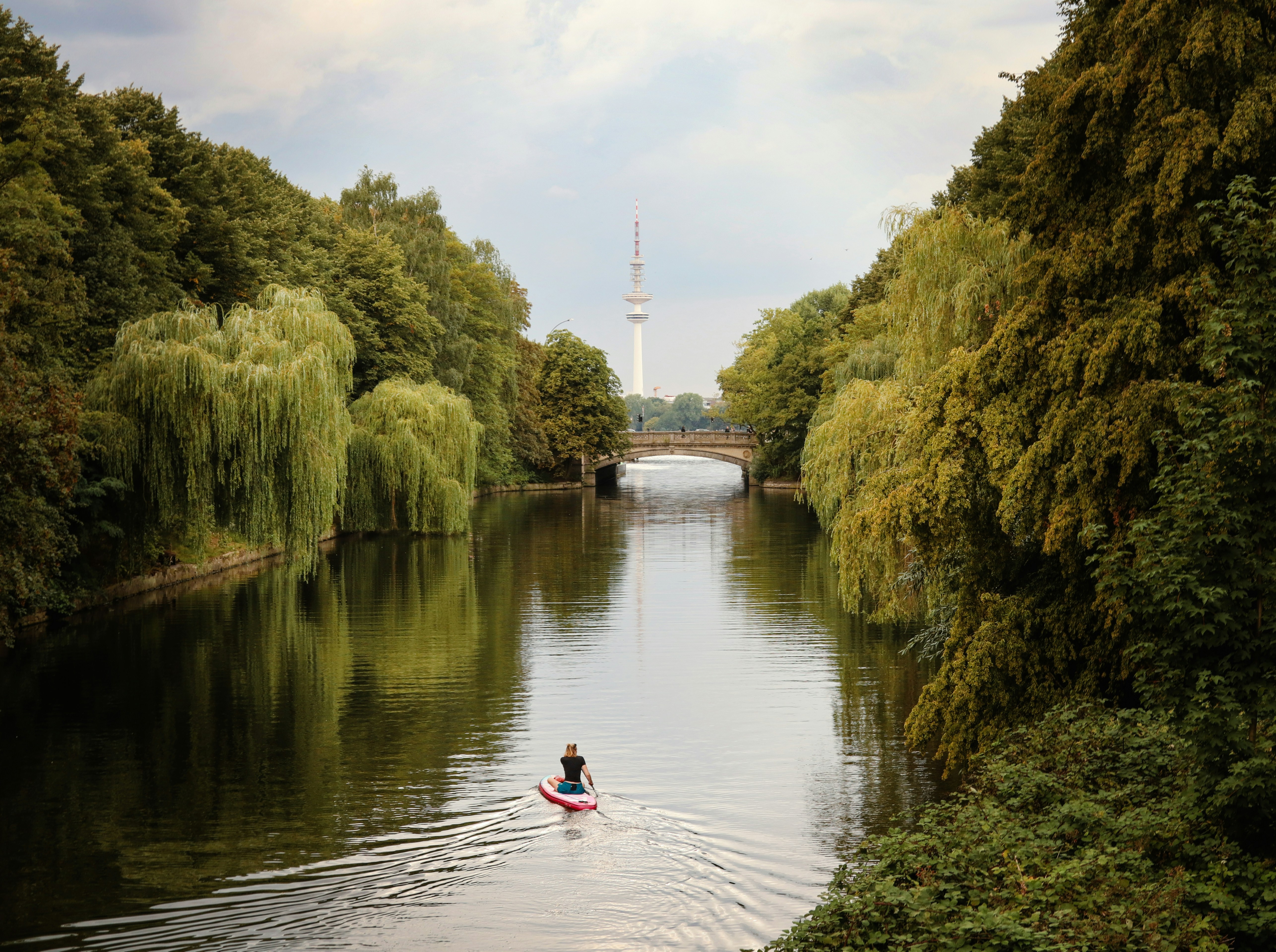 A lone paddler navigates the calm waters of a tree-lined canal, with a distant tower rising above the landscape.