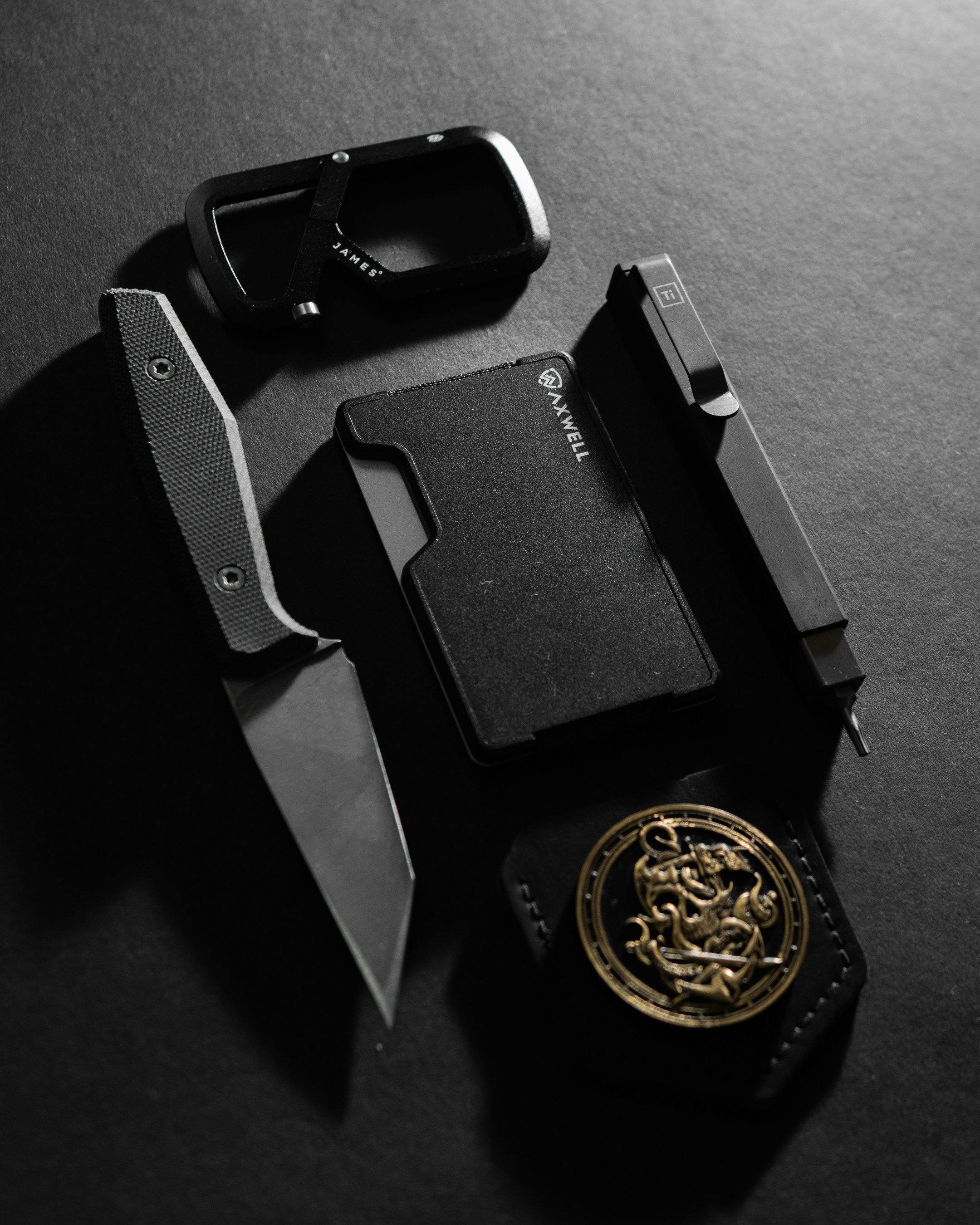 A minimalist arrangement of a knife, wallet, pen, carabiner, and a decorative coin, emphasizing functionality and design against a dark backdrop.