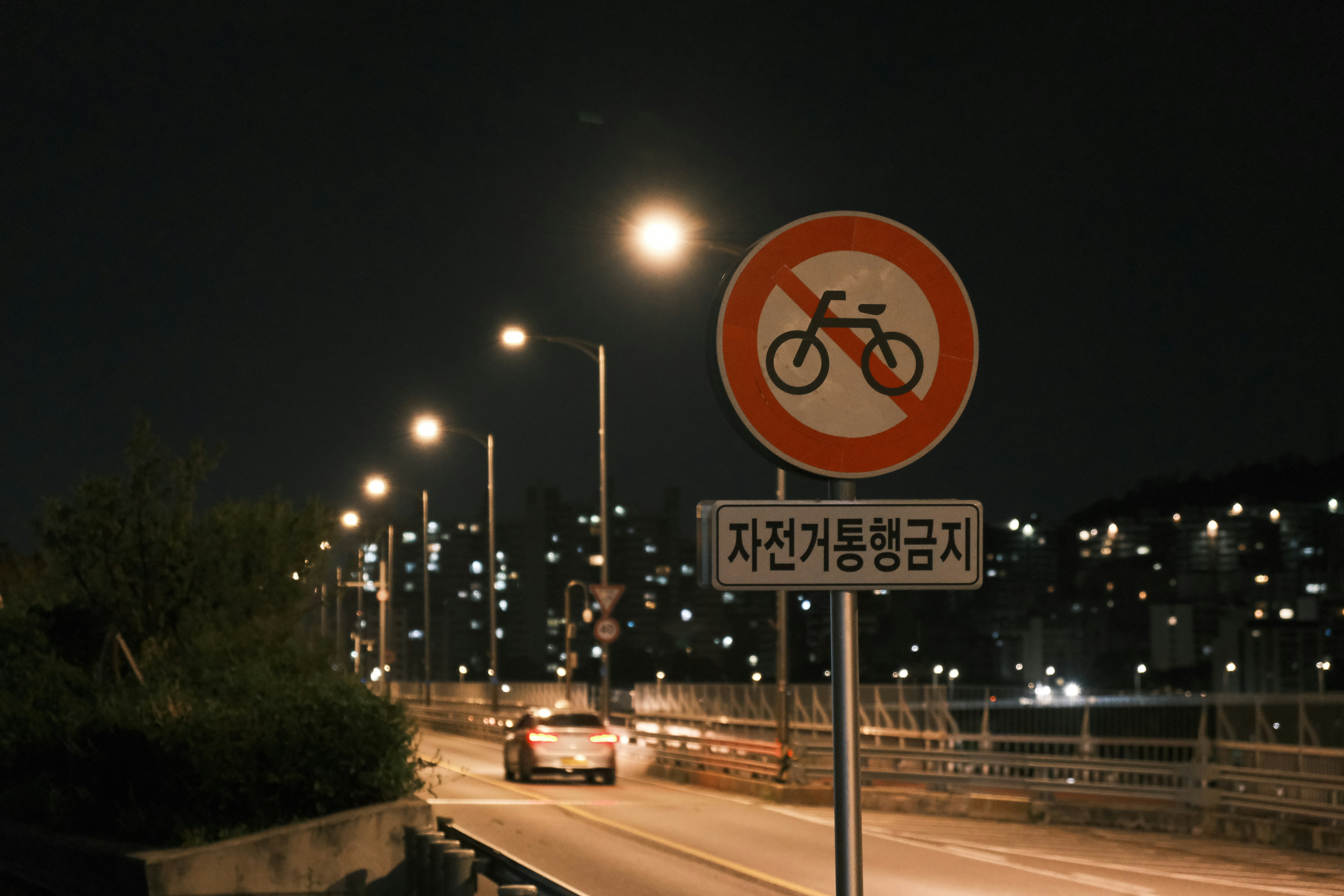 No bicycles sign illuminated by streetlights on a quiet night road with distant city lights.