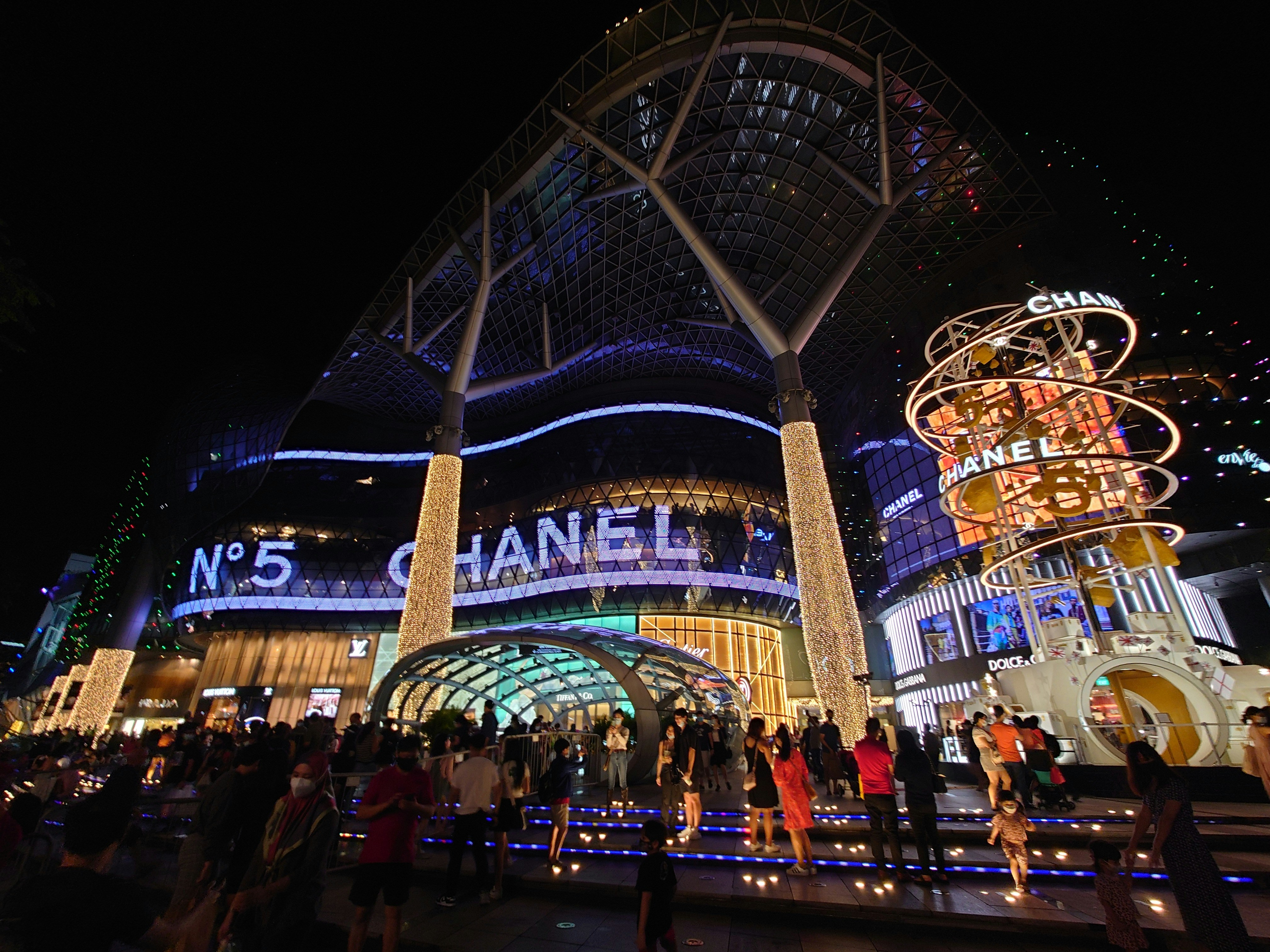 Crowd gathers under vibrant lights at a modern shopping complex at night.
