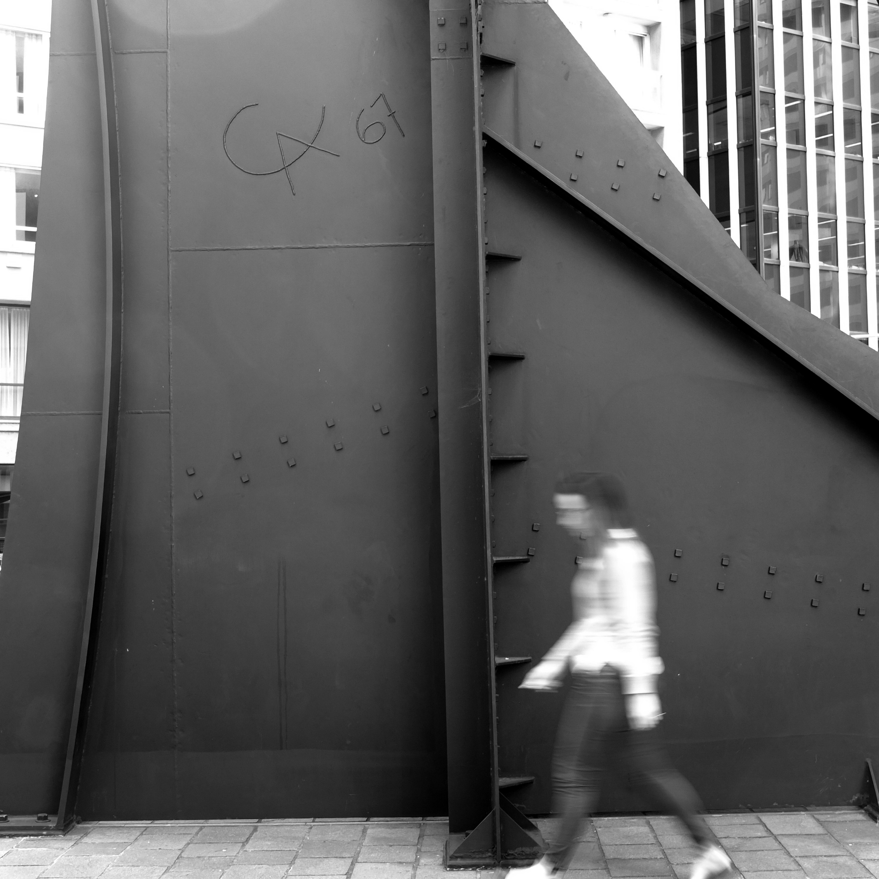 Grayscale photograph of a blurred pedestrian walking past riveted steel panels and diagonal braces.
