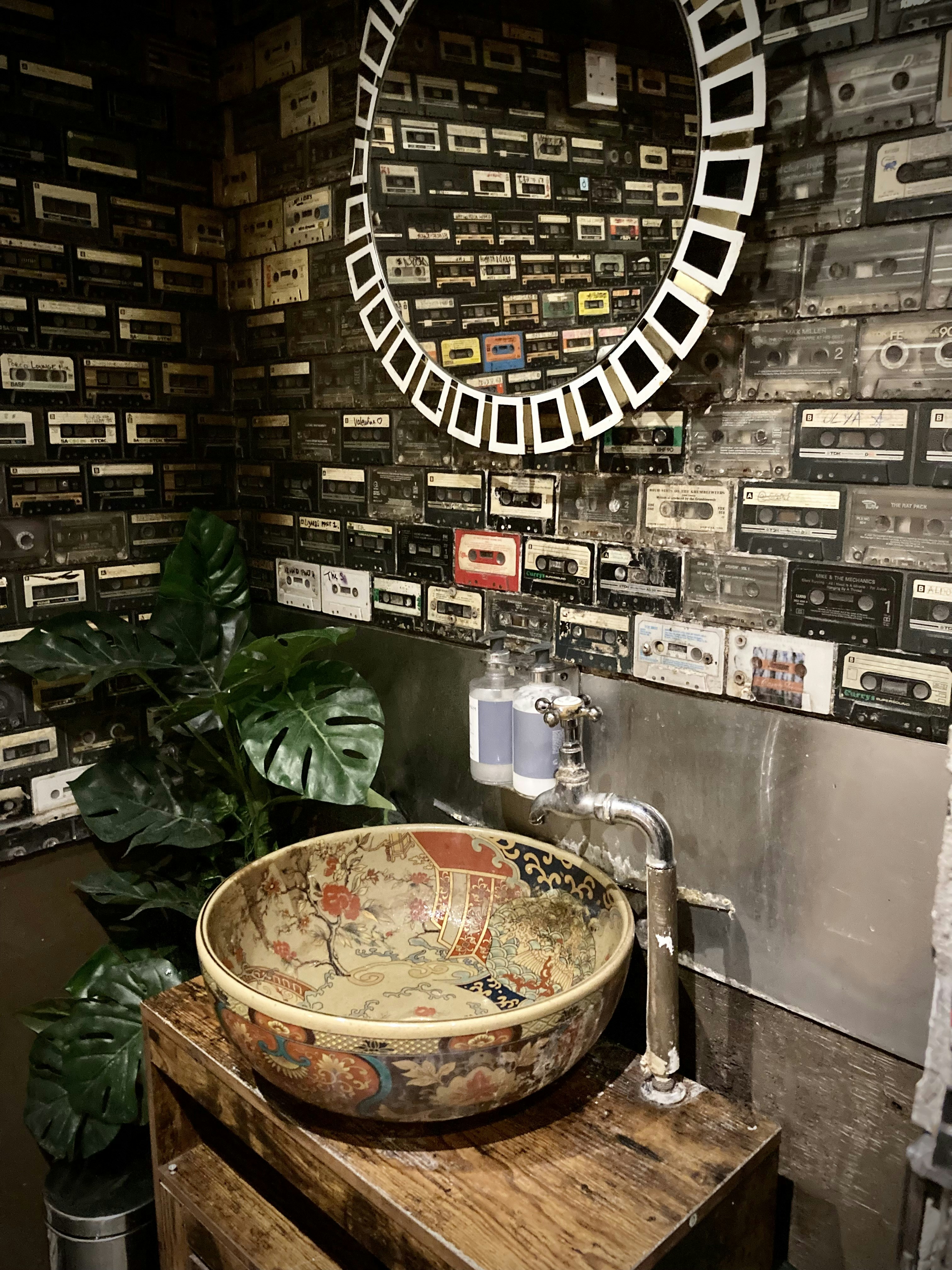 Colorful ceramic basin on a weathered pedestal beneath a circular mirror, against a cassette-tape wallpaper backdrop