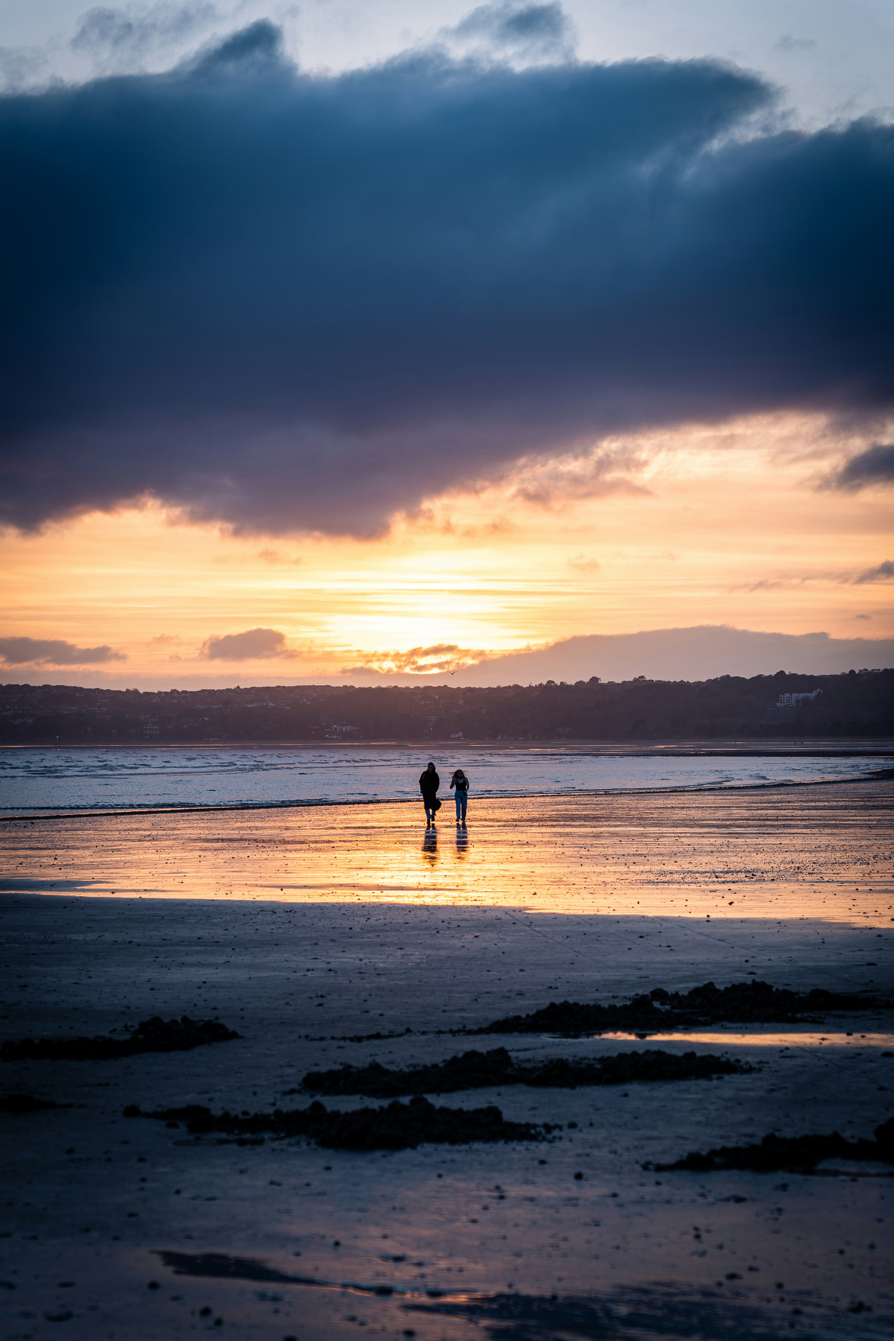 Silhouetted figures walking hand in hand on a reflective beach at sunset, with dramatic clouds overhead.