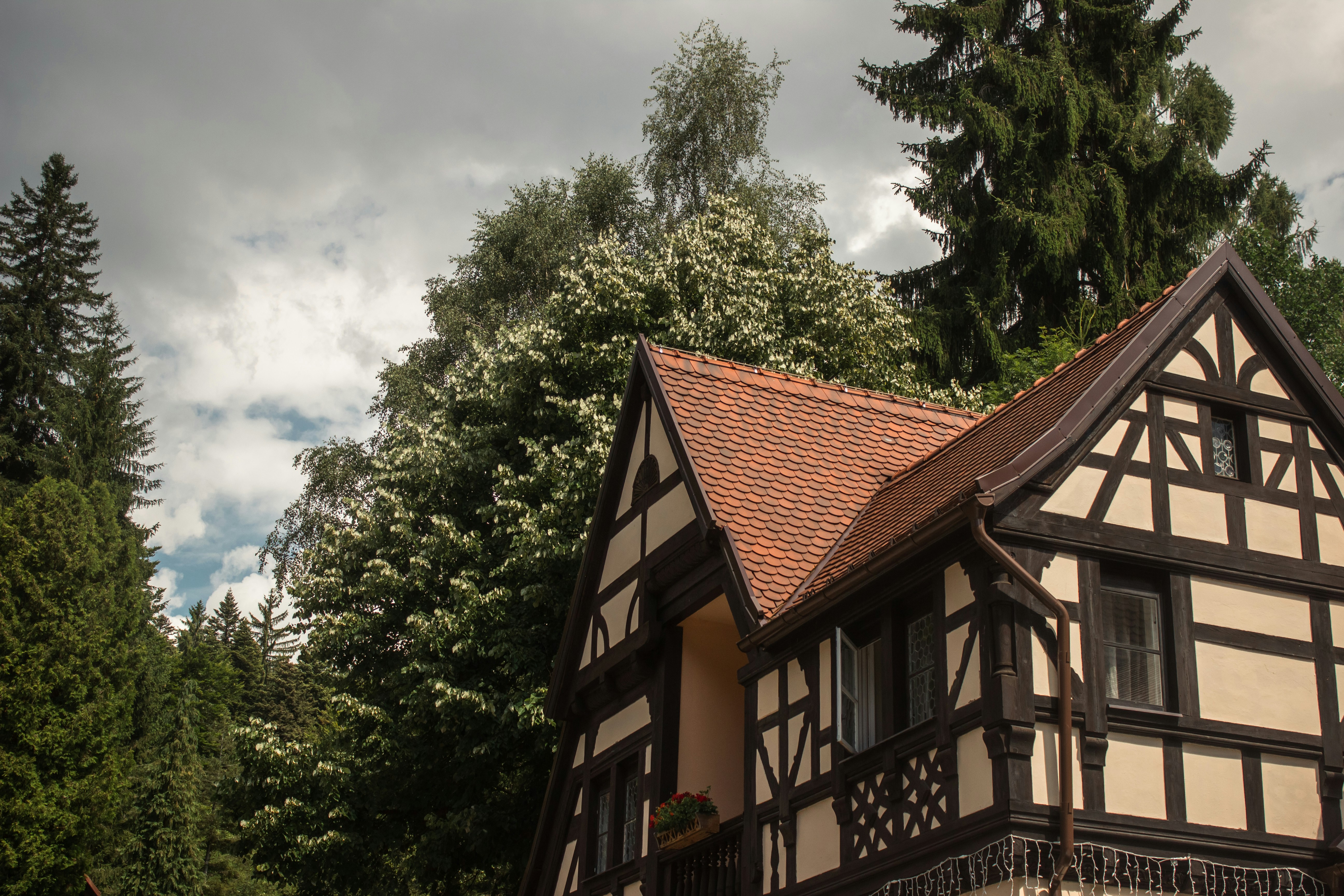 Traditional half-timbered house with a red roof set against a backdrop of tall, lush trees under a cloudy sky.