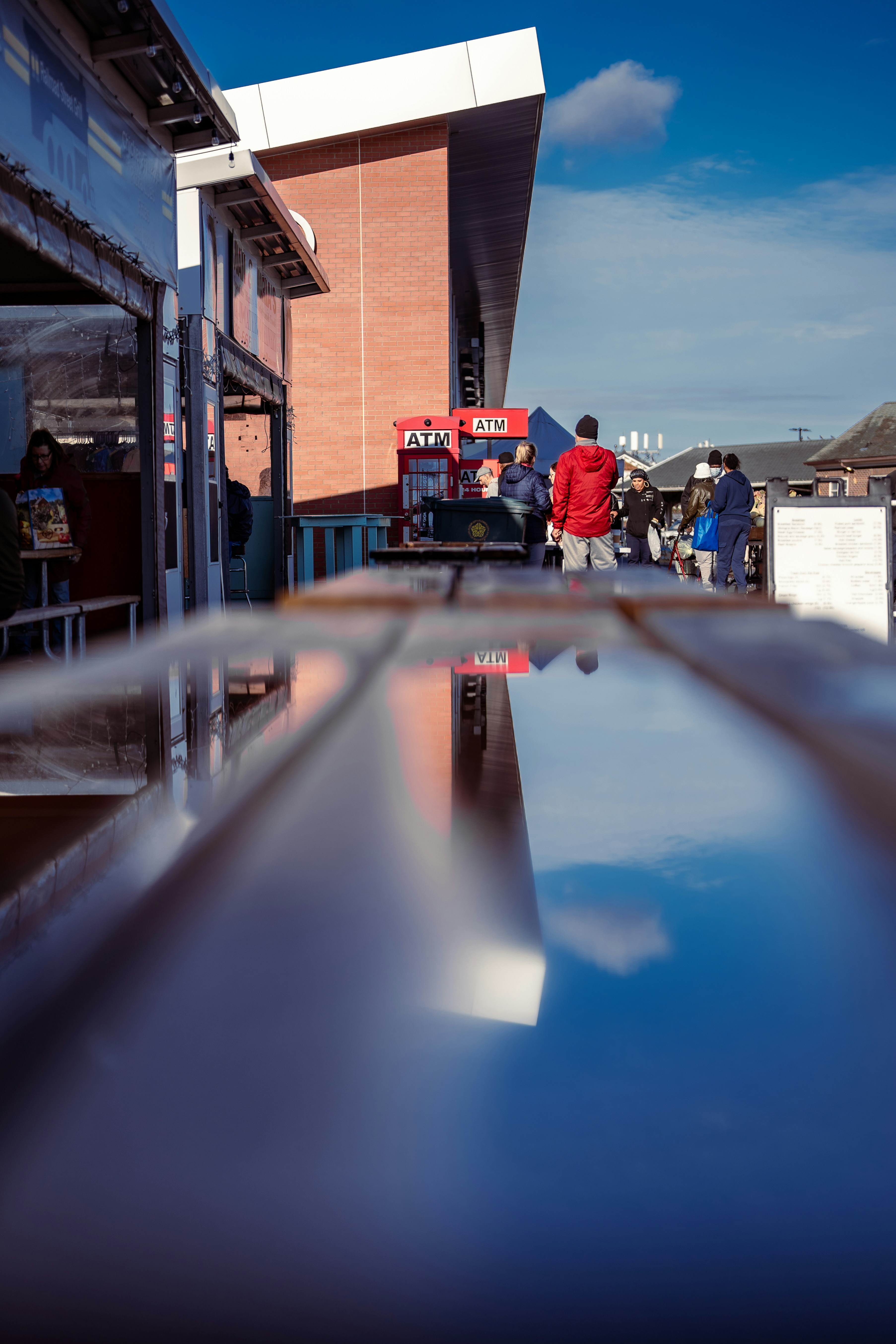 A bustling outdoor scene featuring people interacting near an ATM, with reflections captured on a polished surface in the foreground.