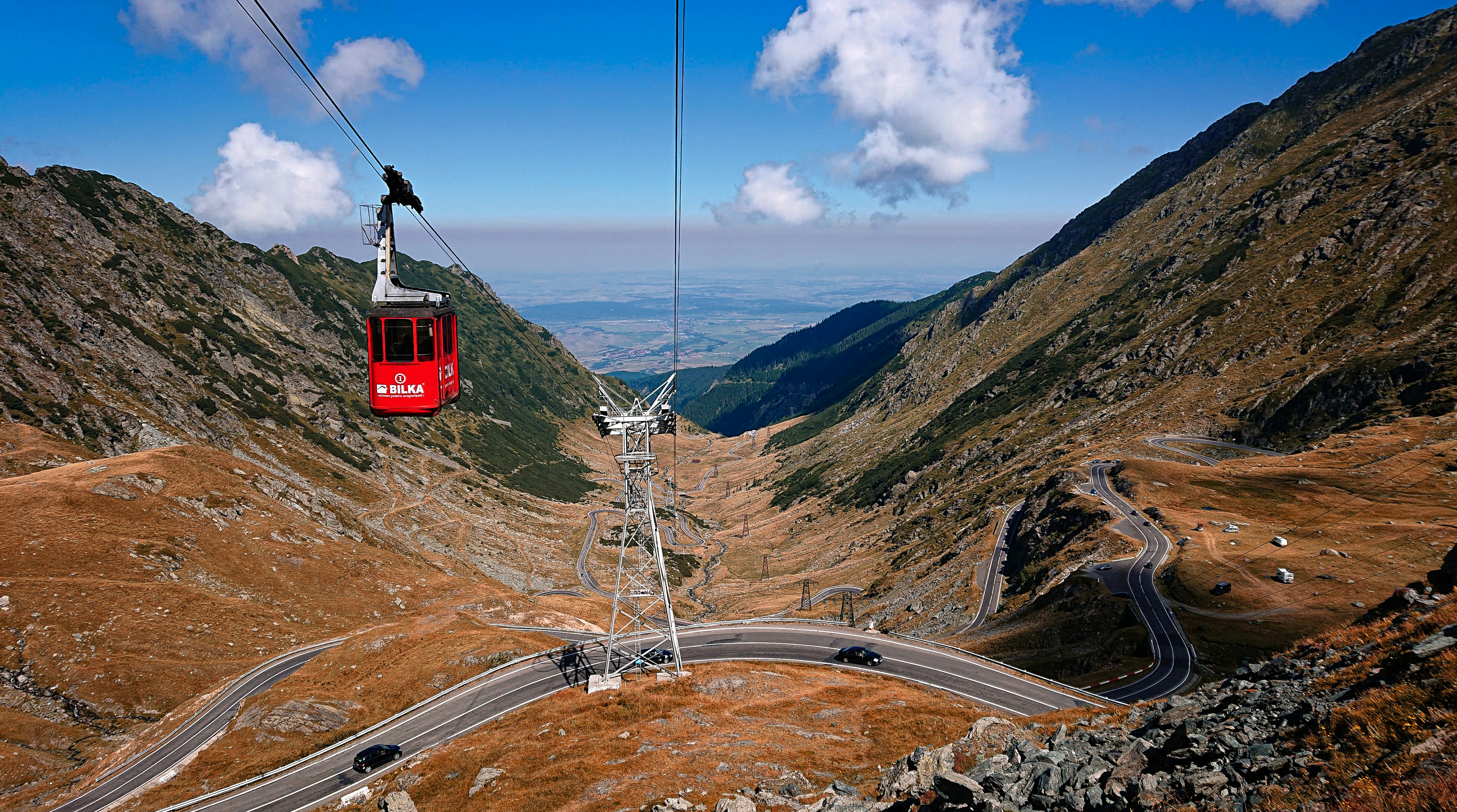Red cable car climbs above a winding mountain road through a rugged valley.