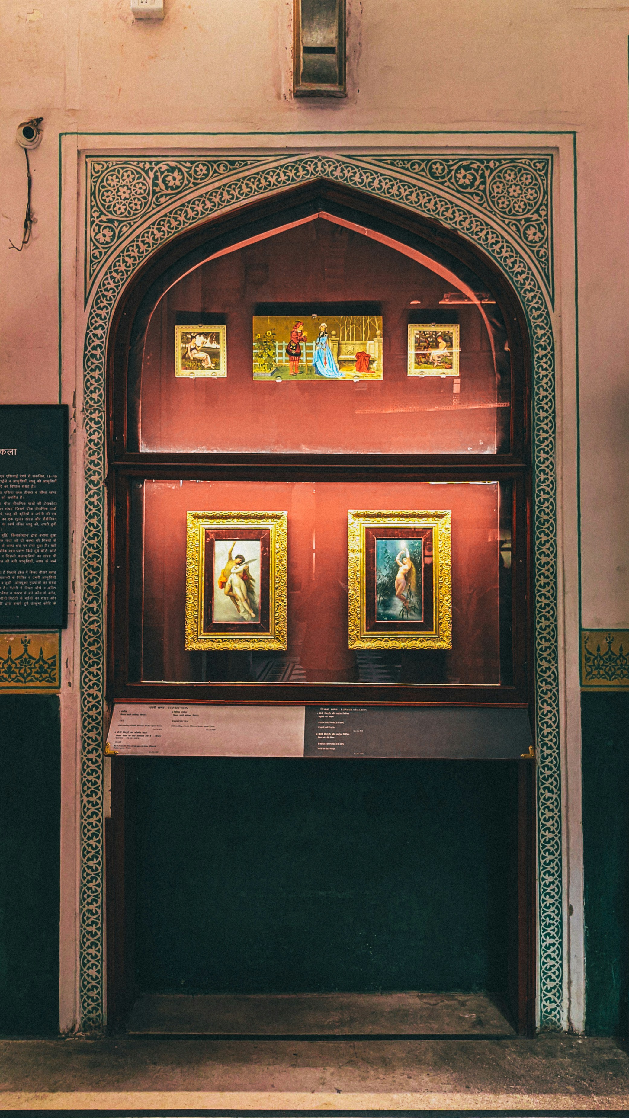 Ornate arched display case on a pink wall with glass, featuring two gold-framed portraits beneath and a row of smaller works above.