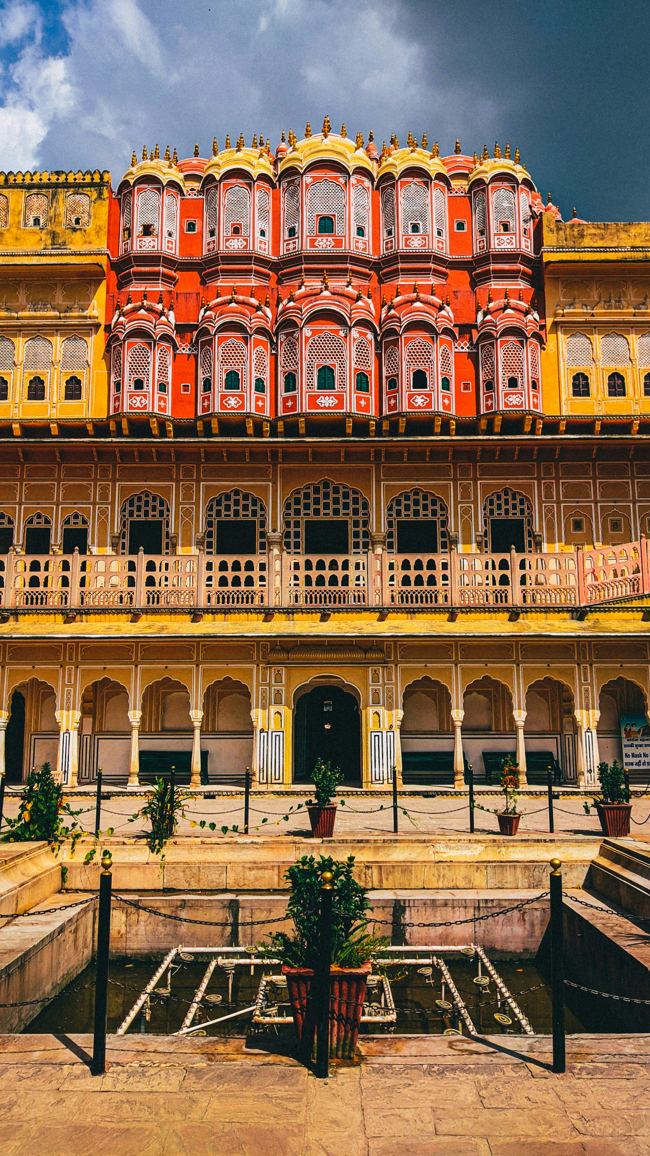 Intricate facade of a historic palace in Jaipur, showcasing vibrant colors and detailed architecture. Lush greenery and a water feature enhance the scene.