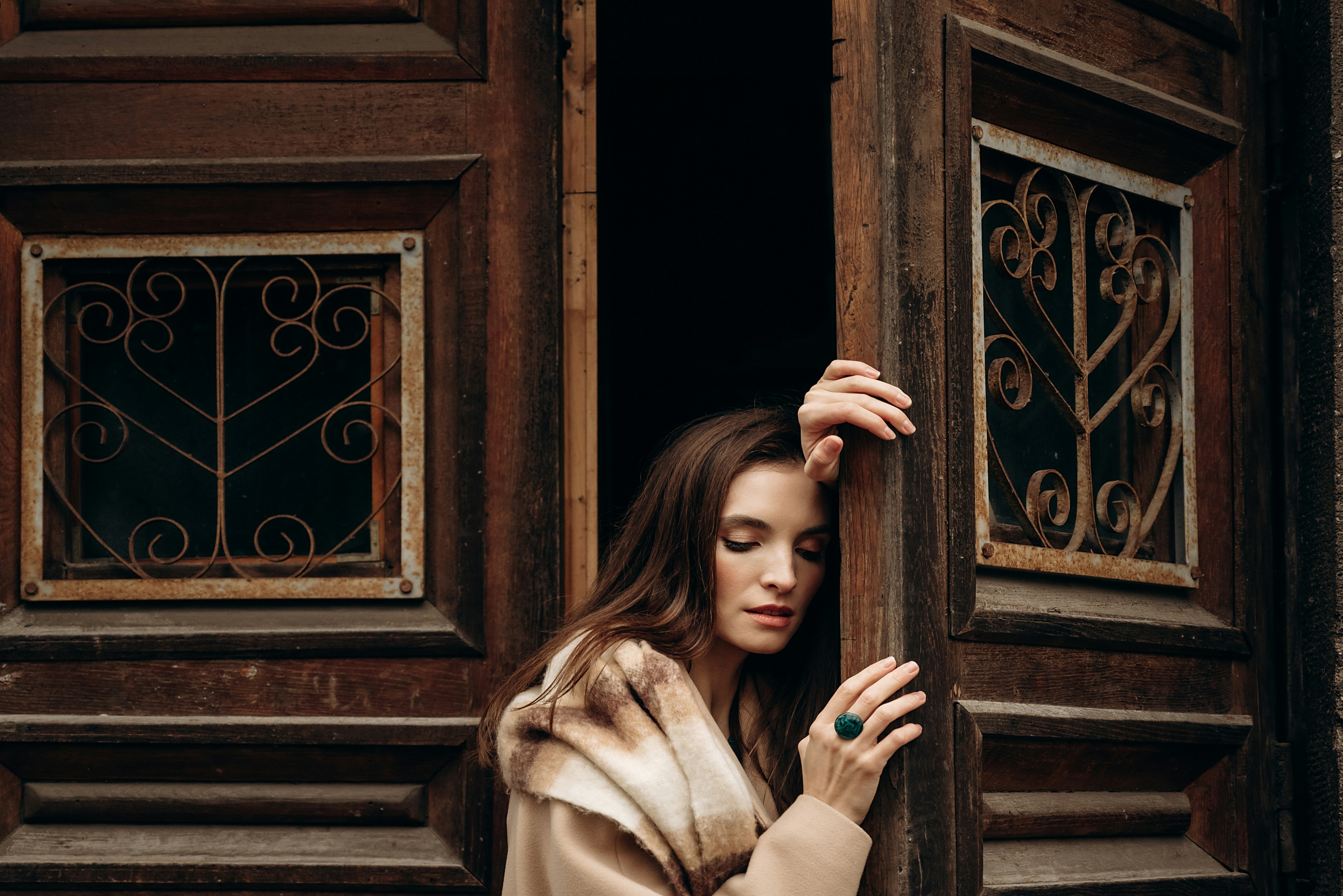 A moody portrait of a woman in a fur-collared coat leaning by a weathered wooden doorway with ornate ironwork.