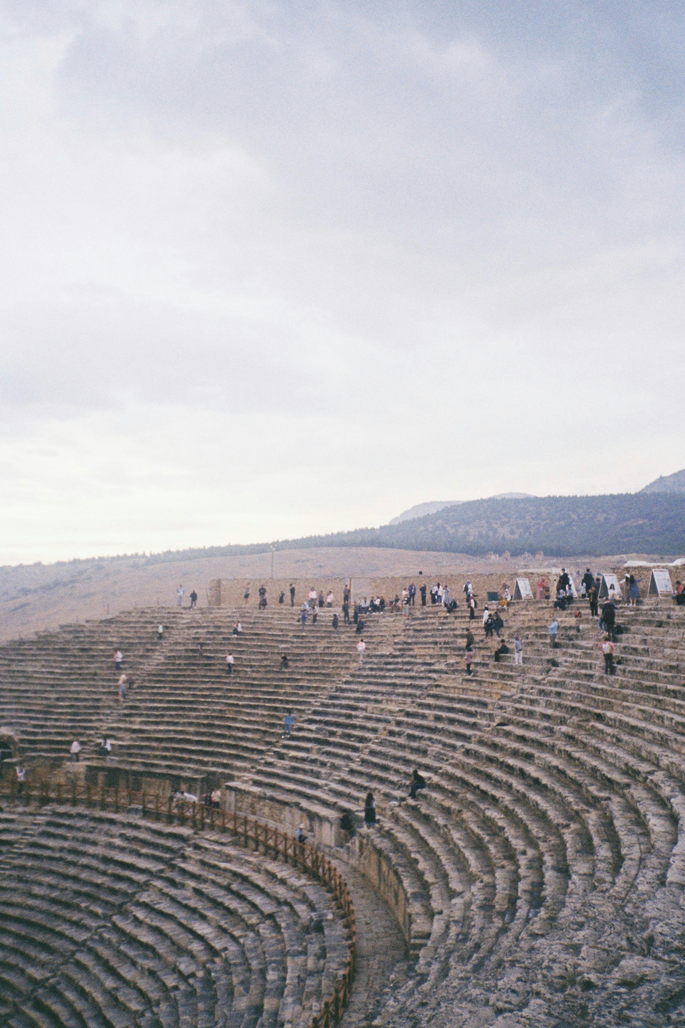 Visitors exploring the tiered seating of an ancient amphitheater under a cloudy sky.