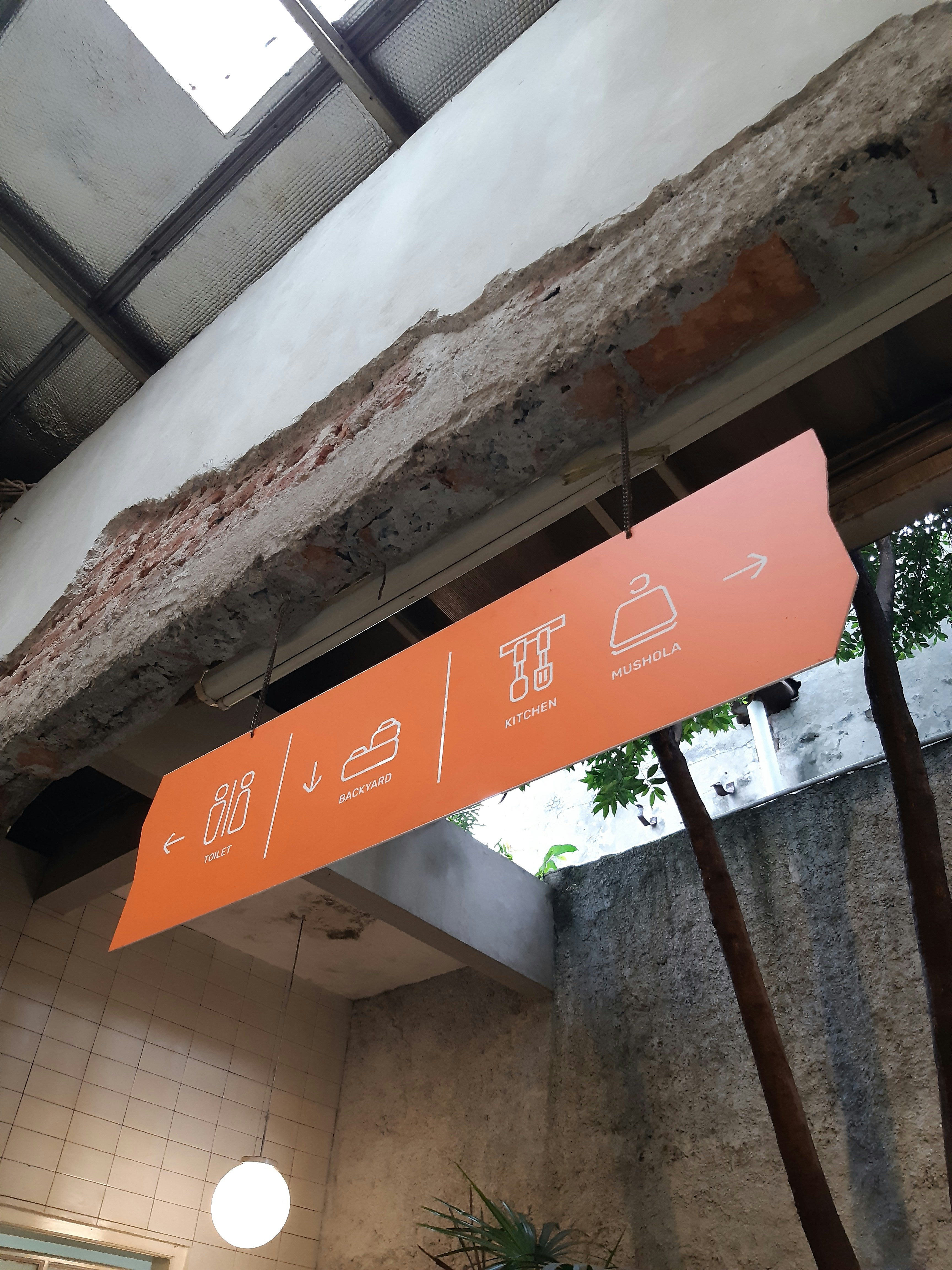 An orange directional signboard hangs from a cracked concrete ceiling, listing toilets, kitchen, and mushola. The interior shows exposed textures and warm, diffused light.