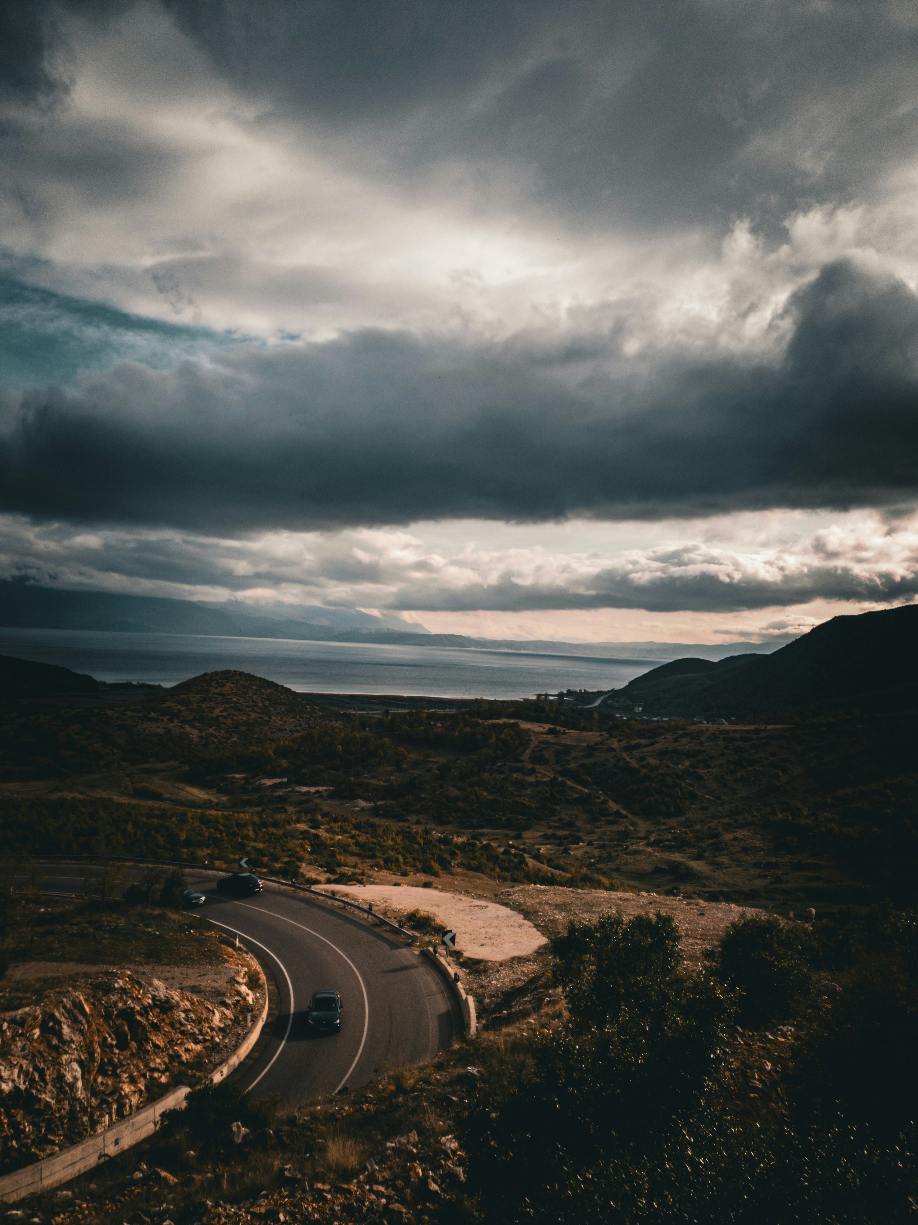 Curved road meandering through a rugged landscape under a dramatic sky, leading towards a distant shoreline.