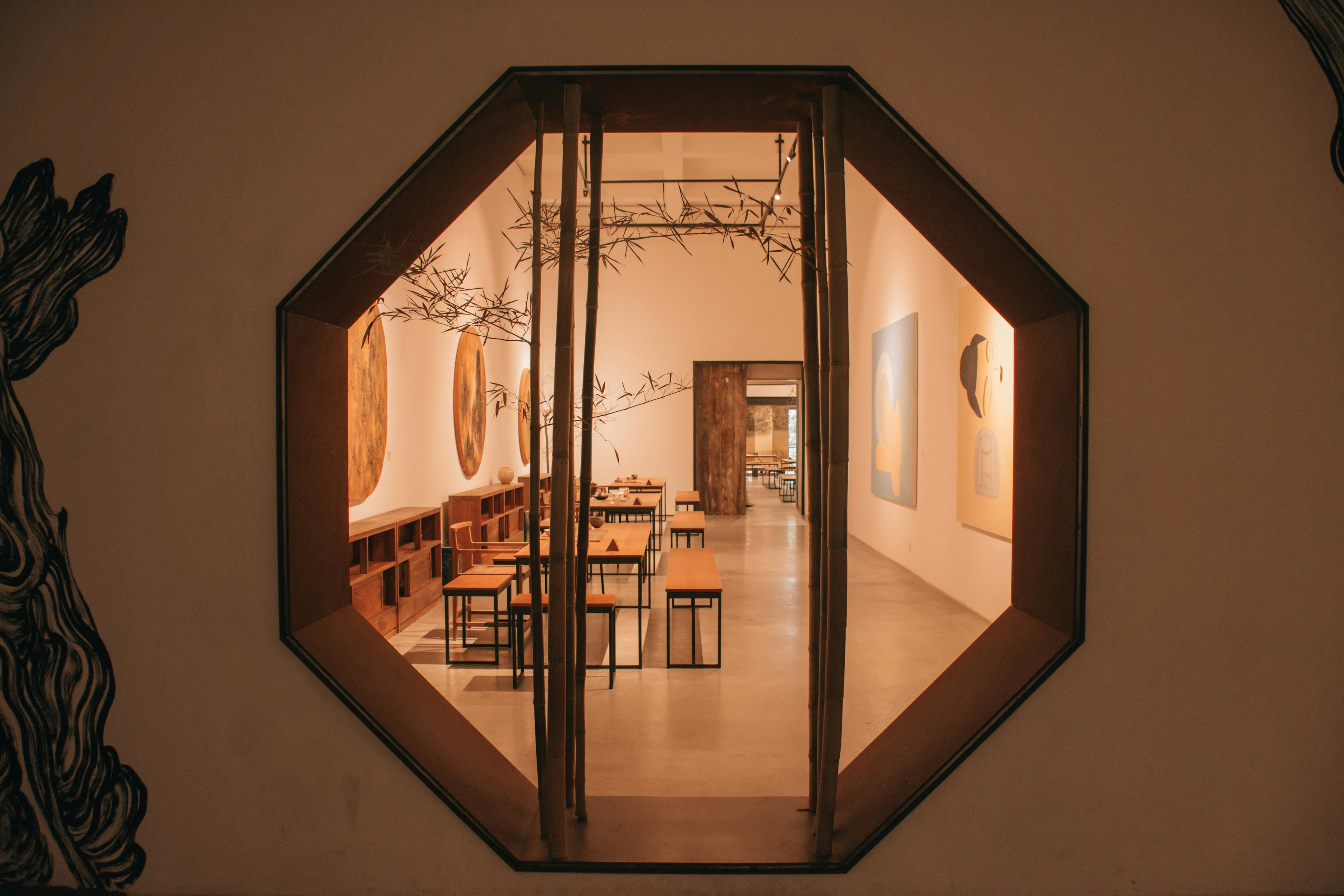 A warm, inviting art studio viewed through an octagonal window, with wooden tables and stools under soft lighting.