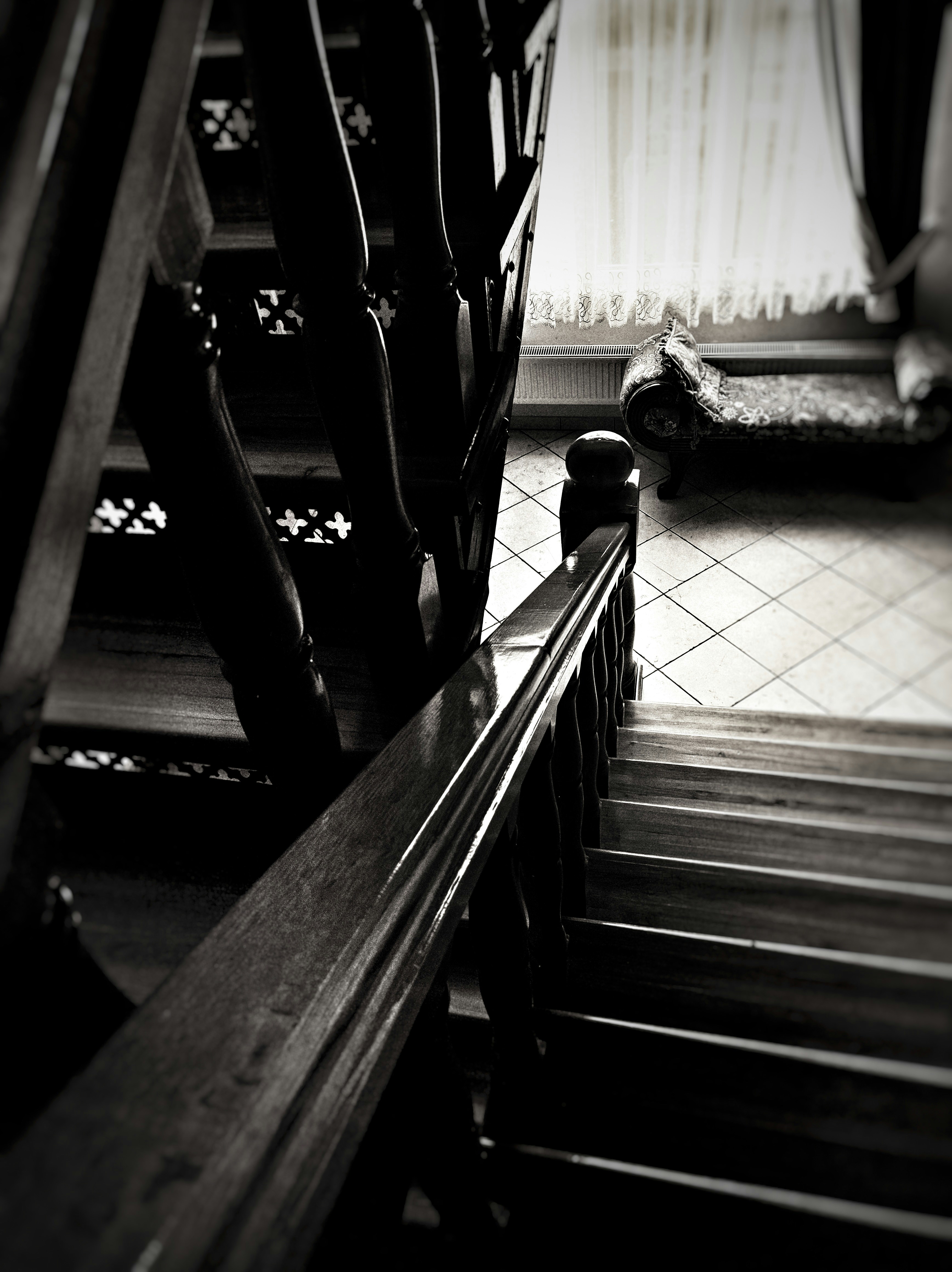 Dim monochrome interior with a wooden staircase and carved balusters; a small silhouette sits by a sunlit window on the landing.