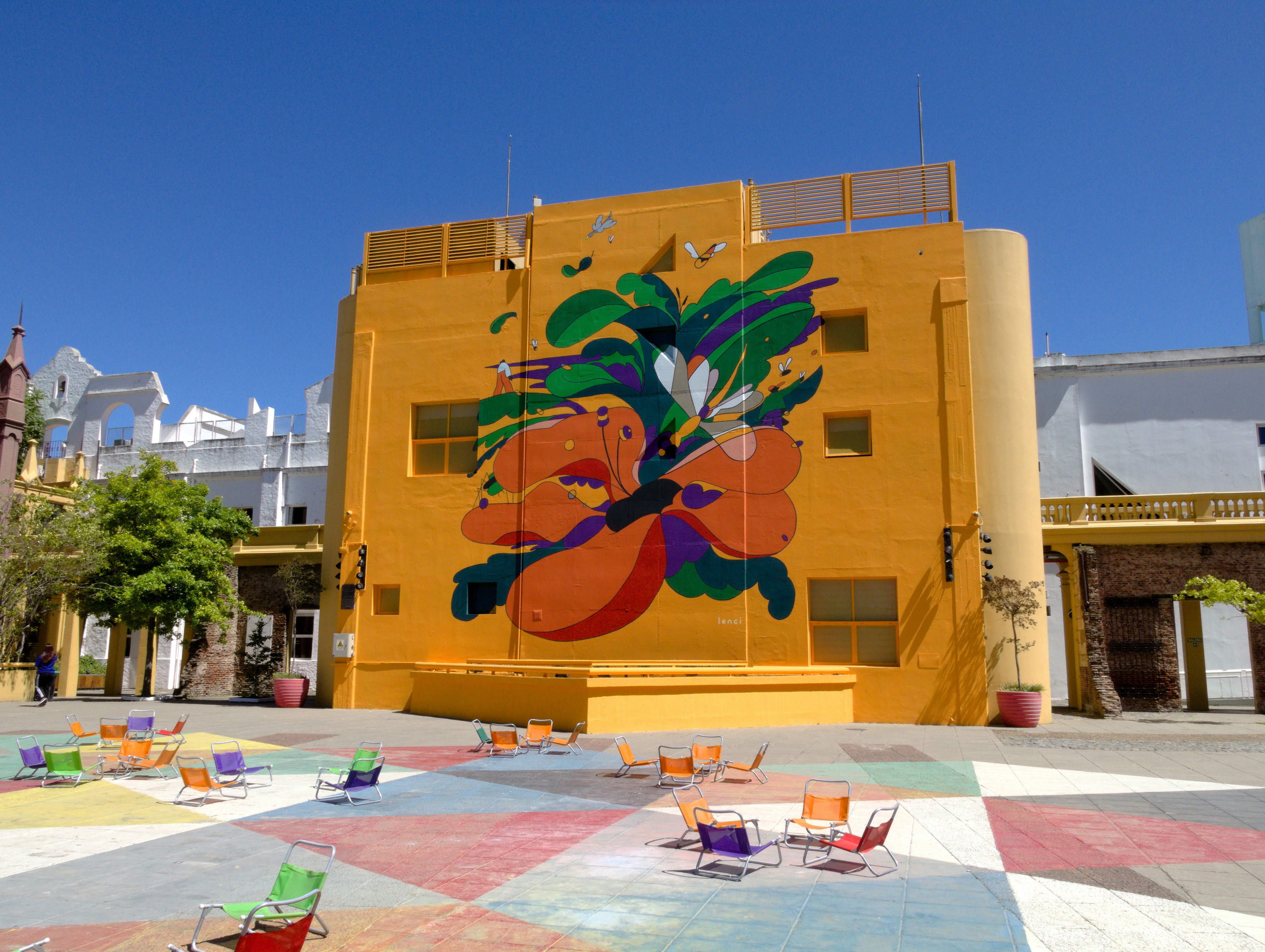 Colorful mural depicting abstract shapes on a bright yellow building under a clear sky.