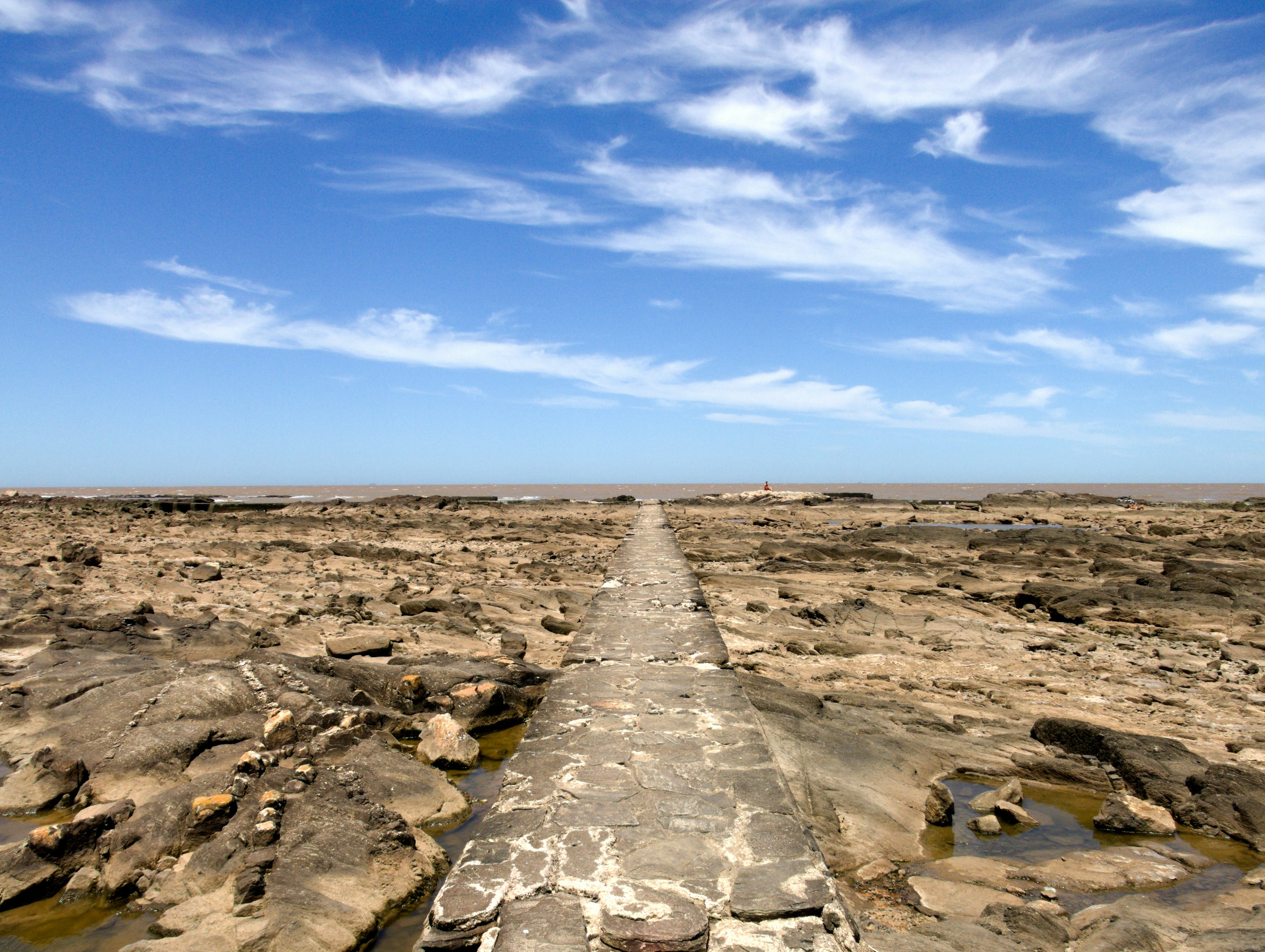 Narrow stone path stretches into a vast, rocky desert under a bright blue sky with scattered clouds.