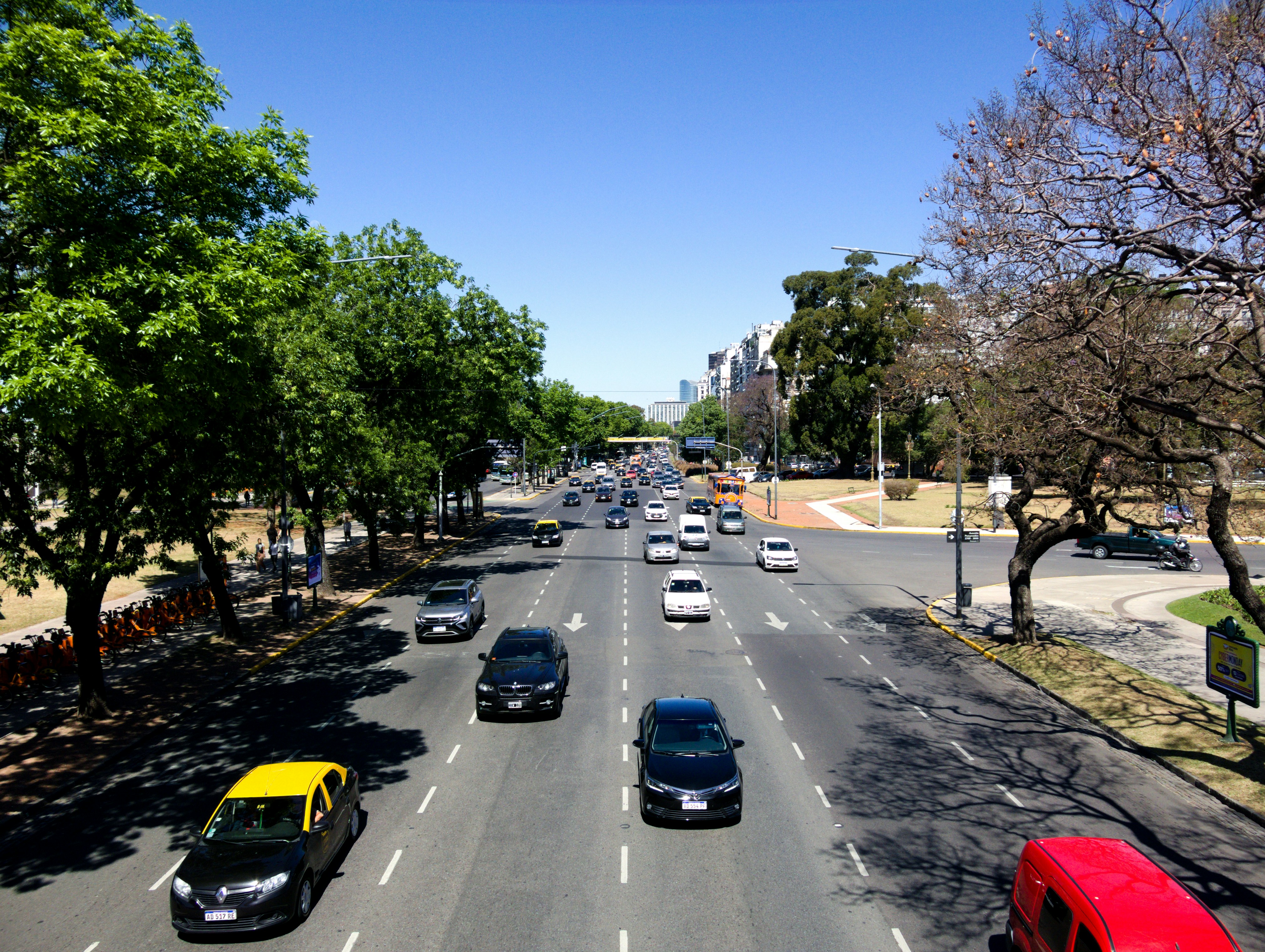Bird's-eye photograph of a busy city avenue, with multiple lanes of traffic flanked by green trees under a clear blue sky.