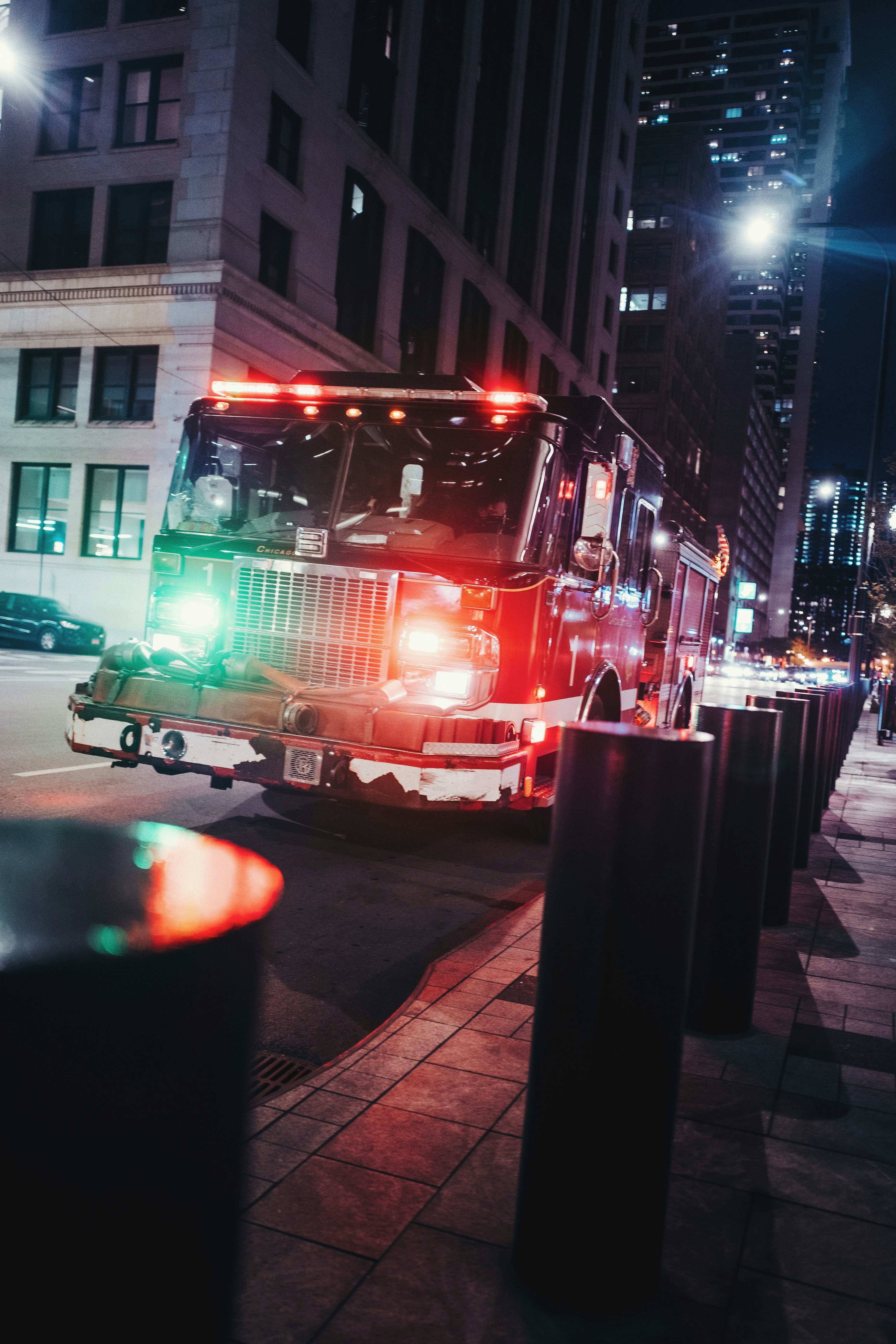 Fire truck parked on a city street at night, illuminated by vibrant emergency lights amidst towering buildings.