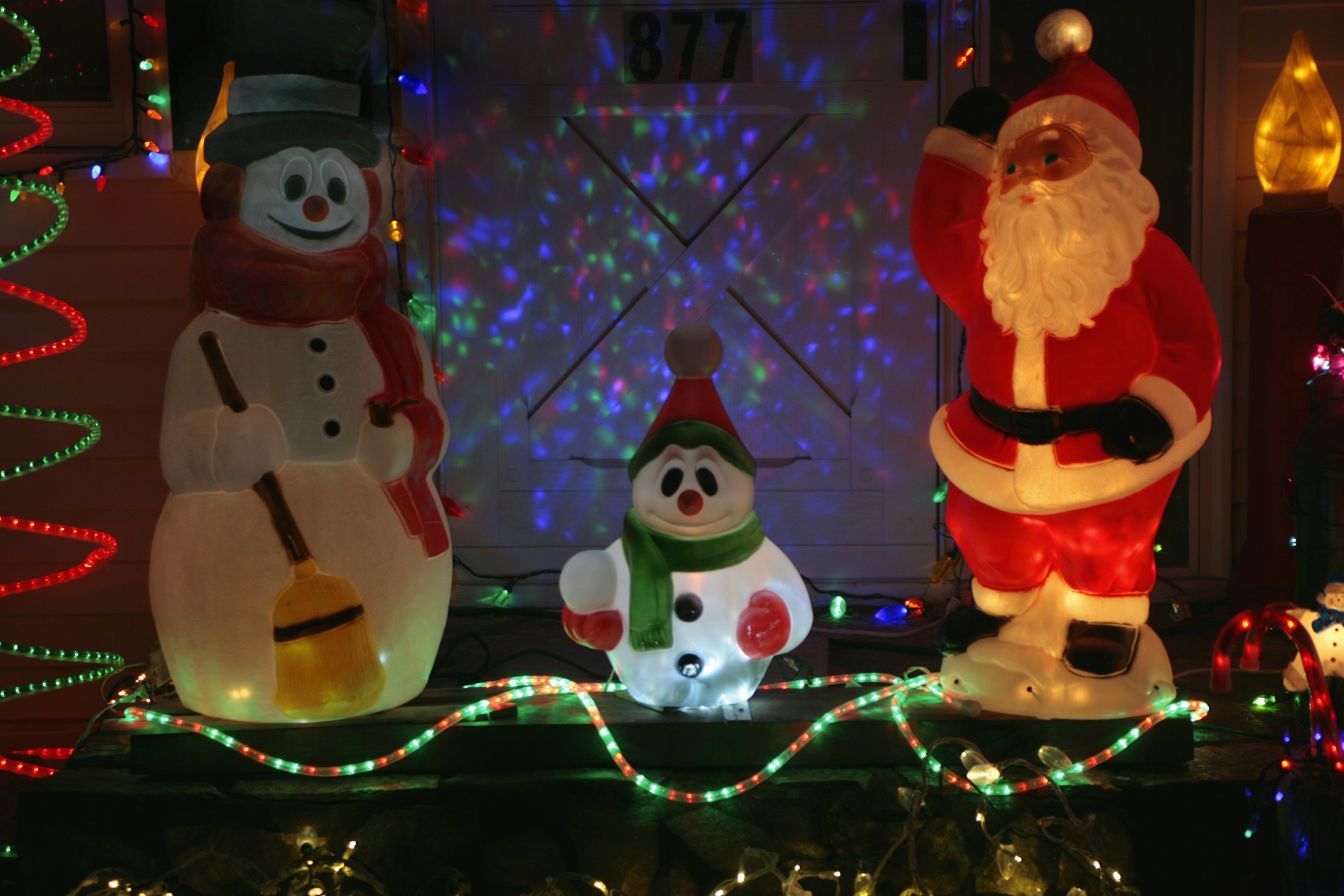 Photograph of inflatable holiday figures—center snowman flanked by a larger snowman on the left and Santa on the right—illuminated by multicolored holiday lights. The scene captures a festive storefront display.