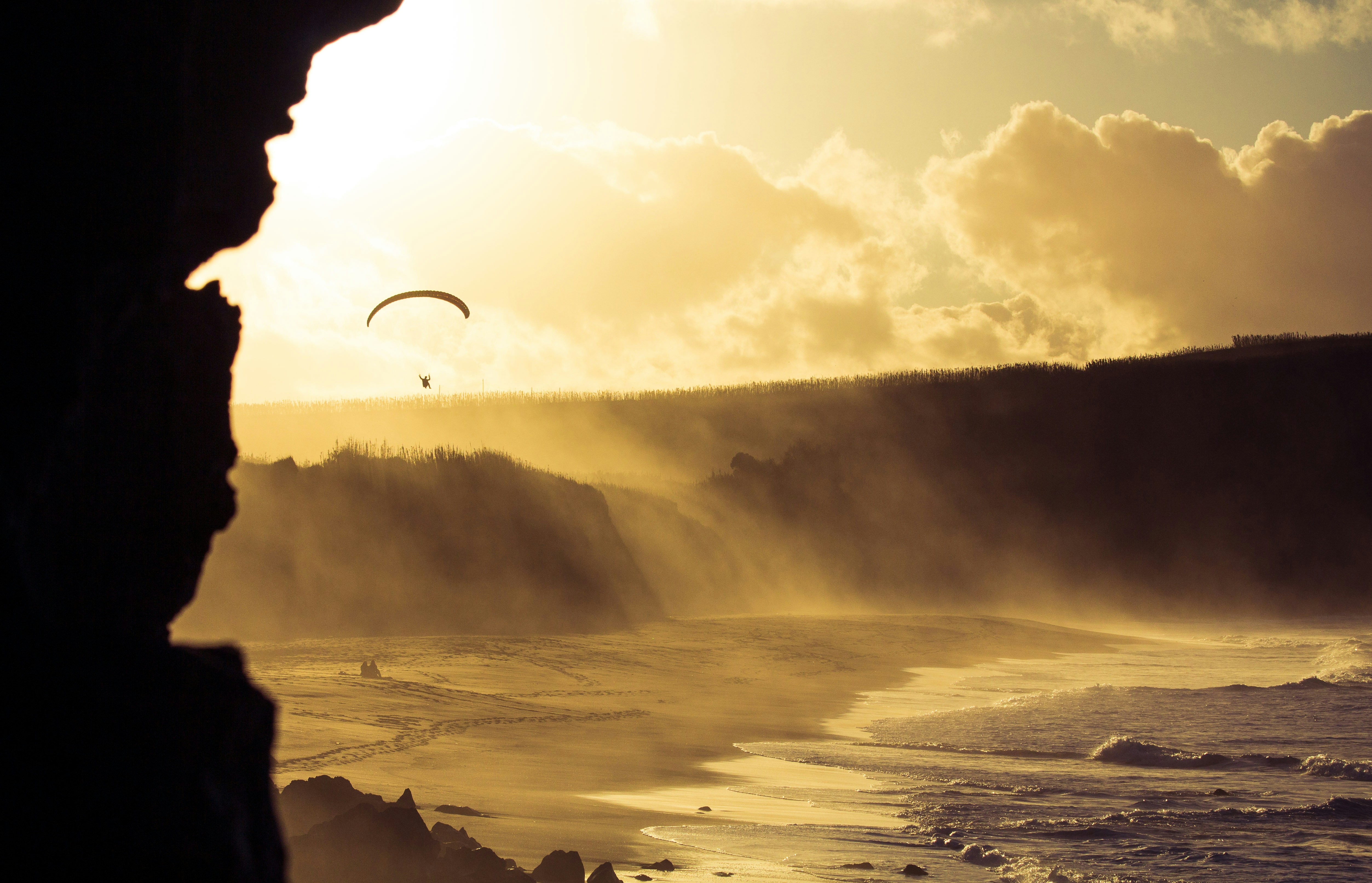 Paraglider navigating above a sunlit beach with mist rising from the waves at sunset.