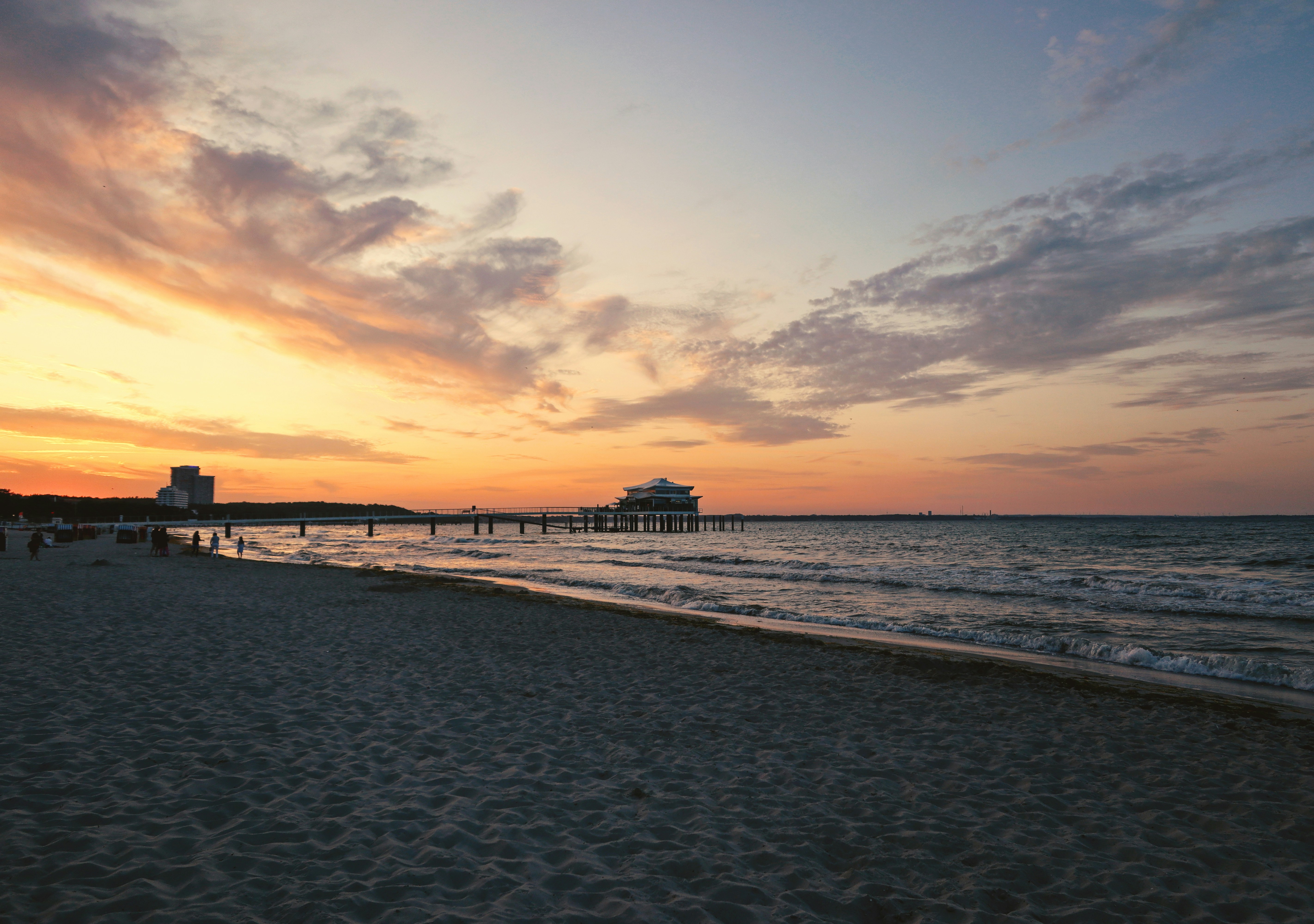 Sunset bathes the beach in warm light as gentle waves wash the shore. A wooden pier extends toward a distant pavilion, carving a calm focal line against the glowing horizon.